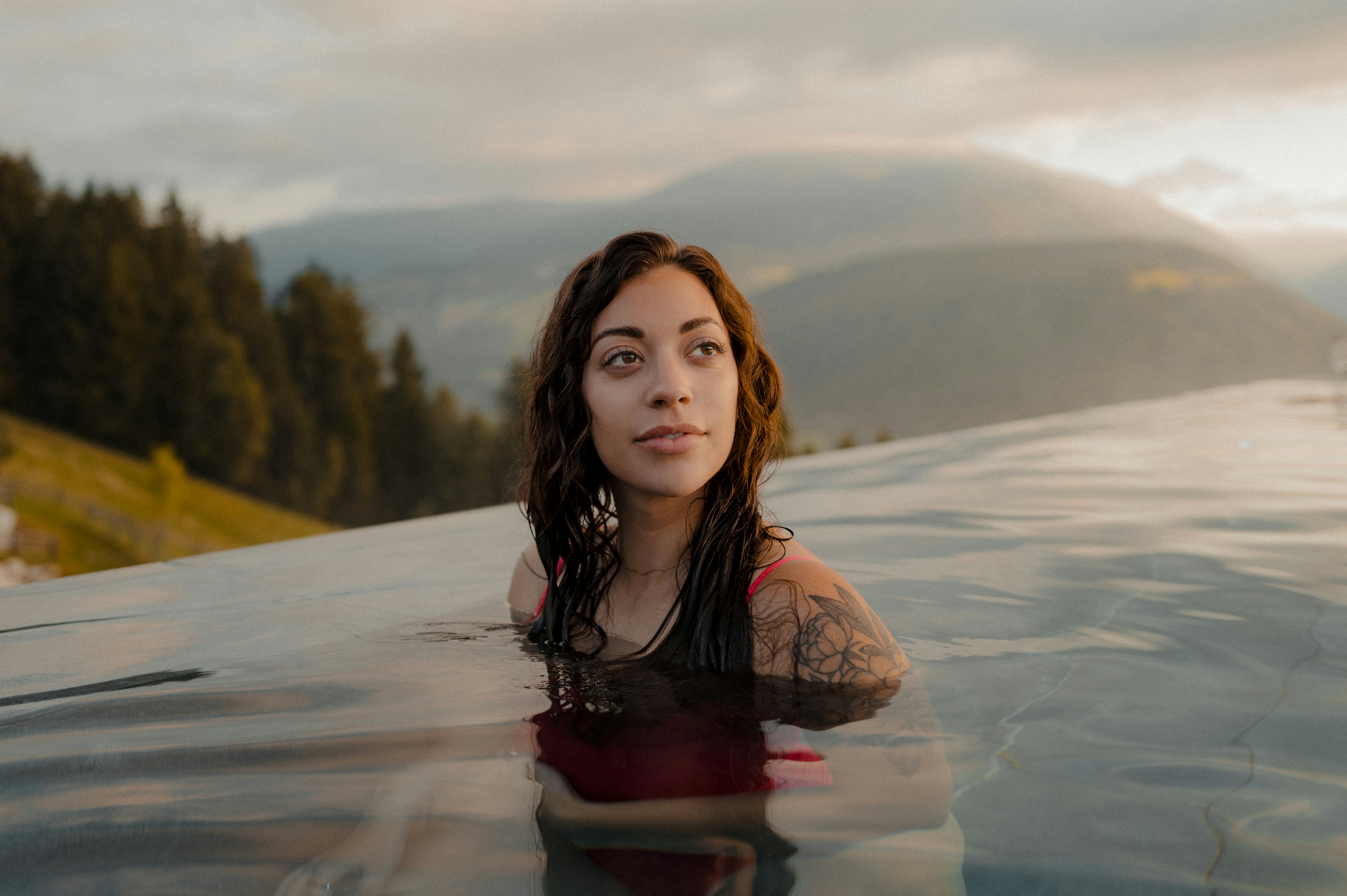 Young woman partially submerged in a pool with mountains and trees in the background, calm expression, warm evening light; a candid portrait emphasizing serenity by the water.