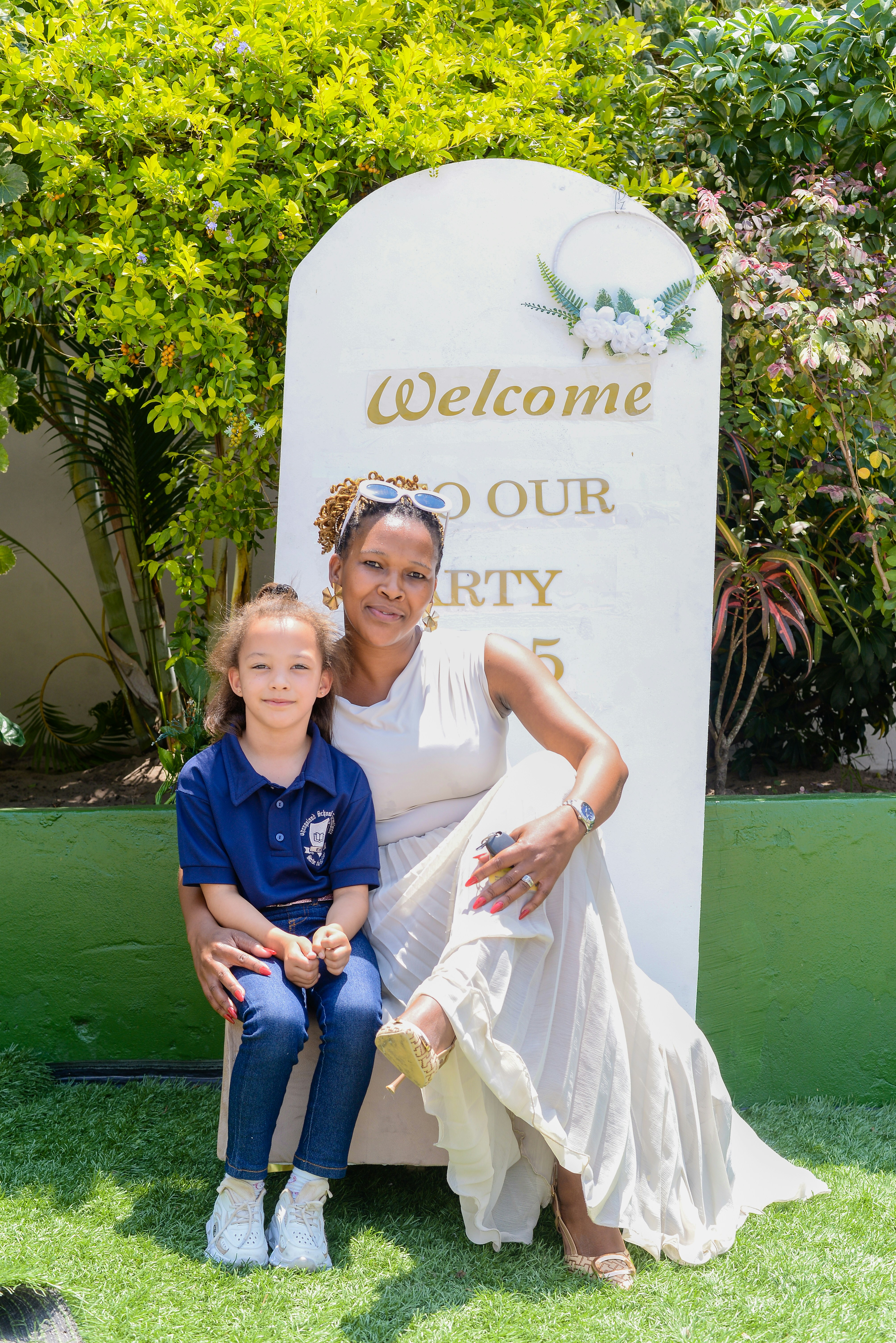 Mother and Daughter Sit Beside a Welcome Sign Under Bright Garden Light