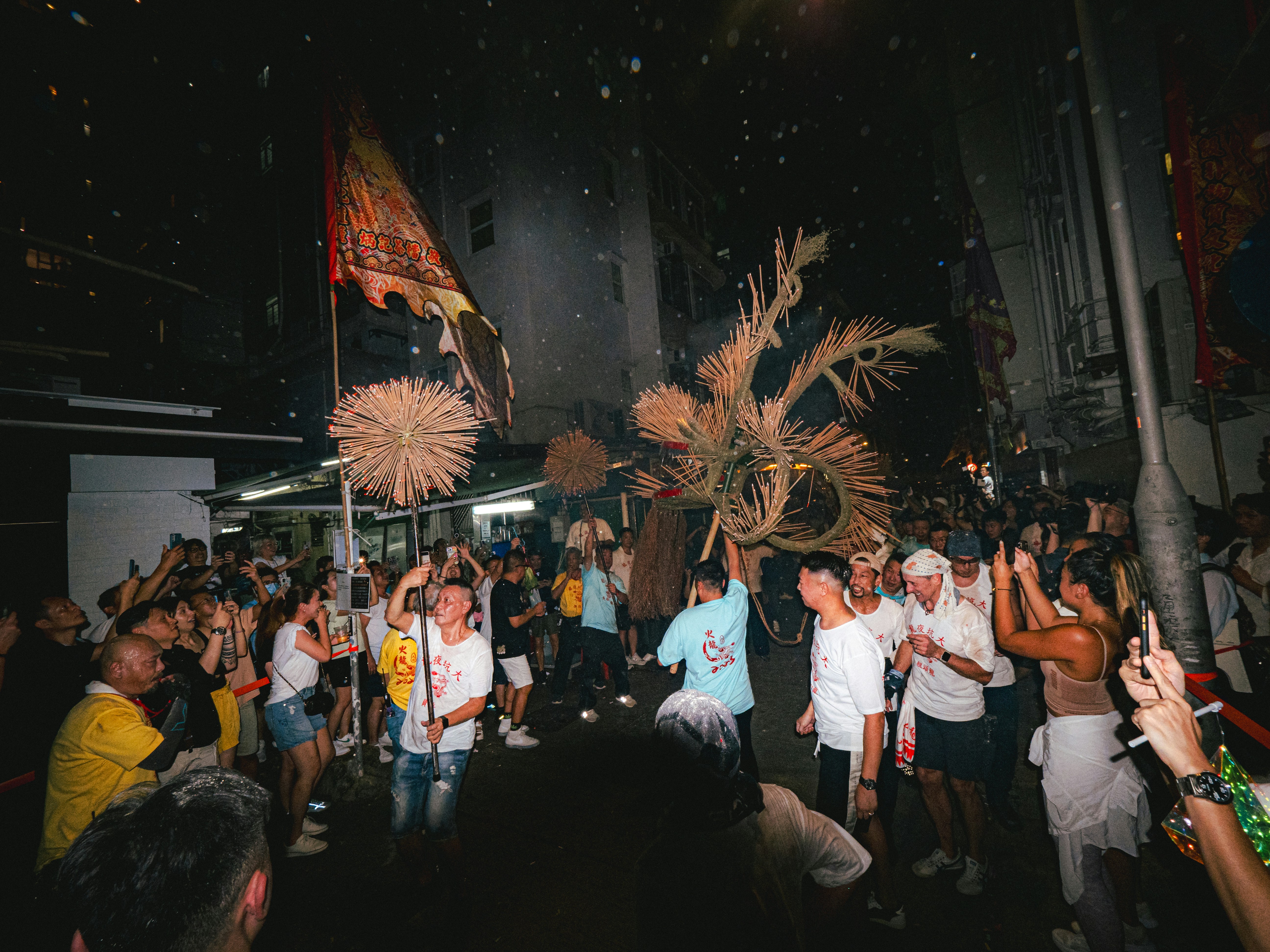 Night street parade with dancers and palm-like banners amid a crowd; palm frond props and umbrellas create a festive performance space.