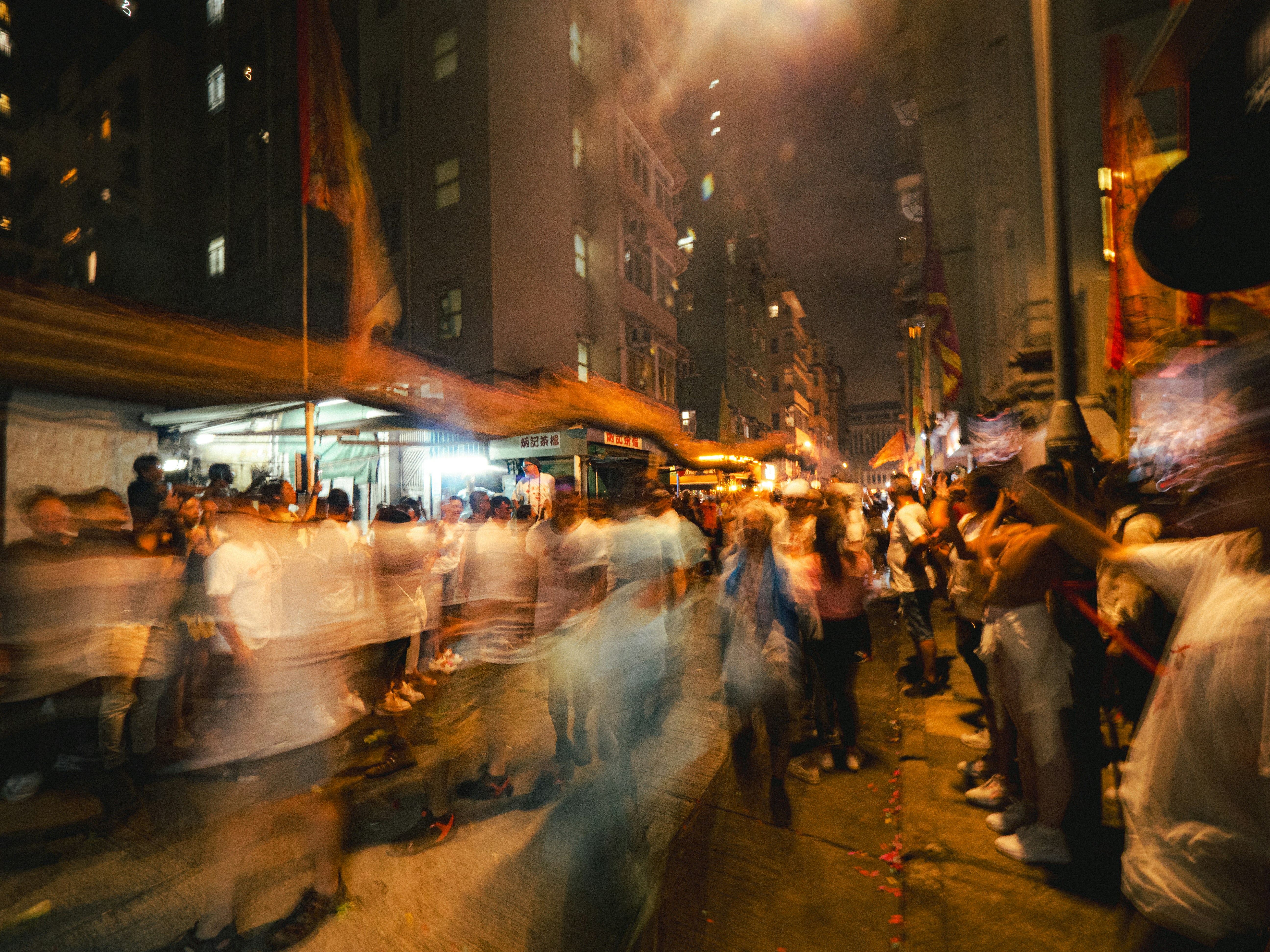 Blurred crowds swirl along a neon-lit street at night, suggesting a bustling festival or market with motionive energy.