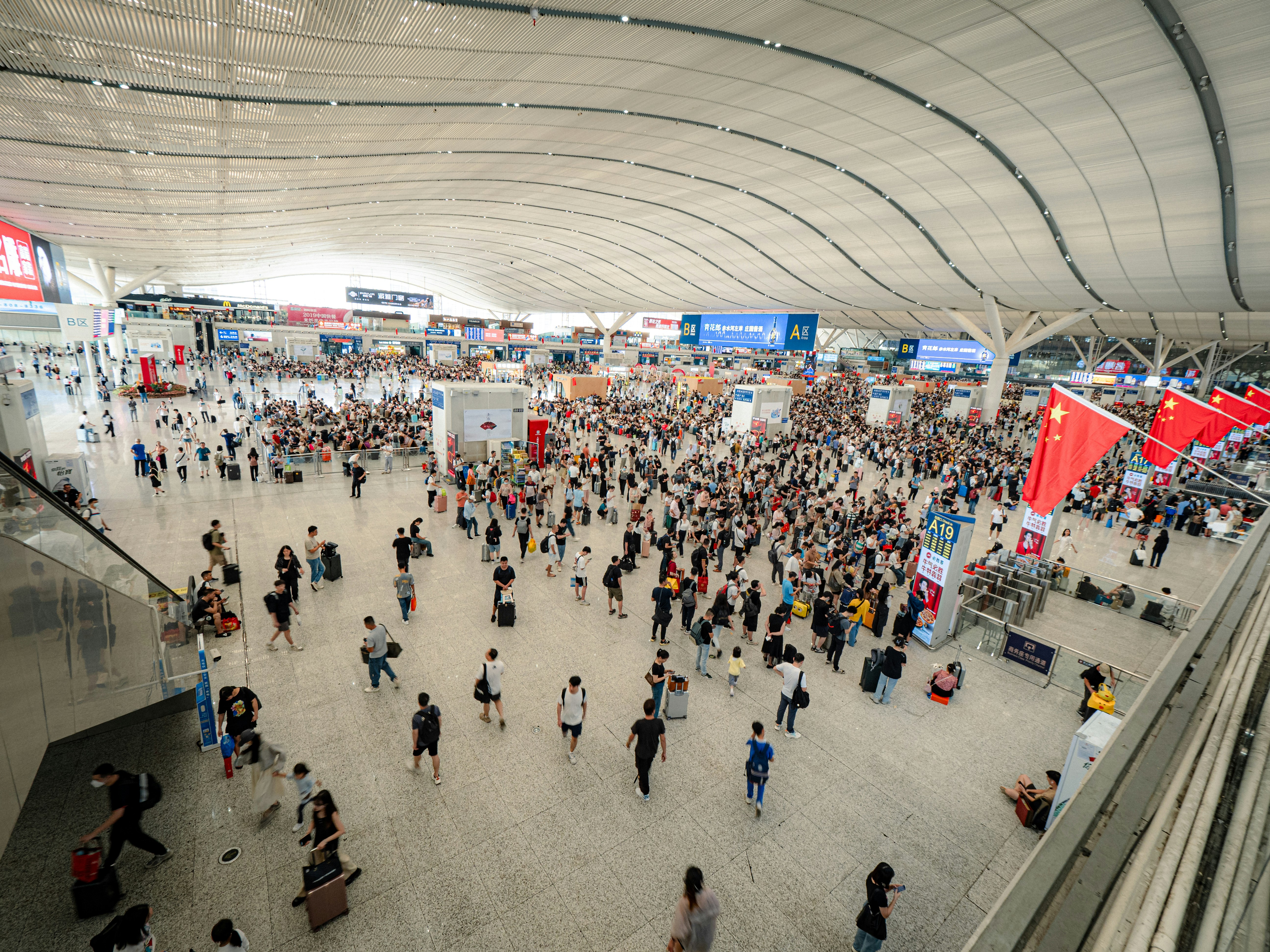 A busy airport-like terminal filled with travelers moving about under a sweeping curved roof; flags and kiosks line the periphery, with a sense of depth and activity.