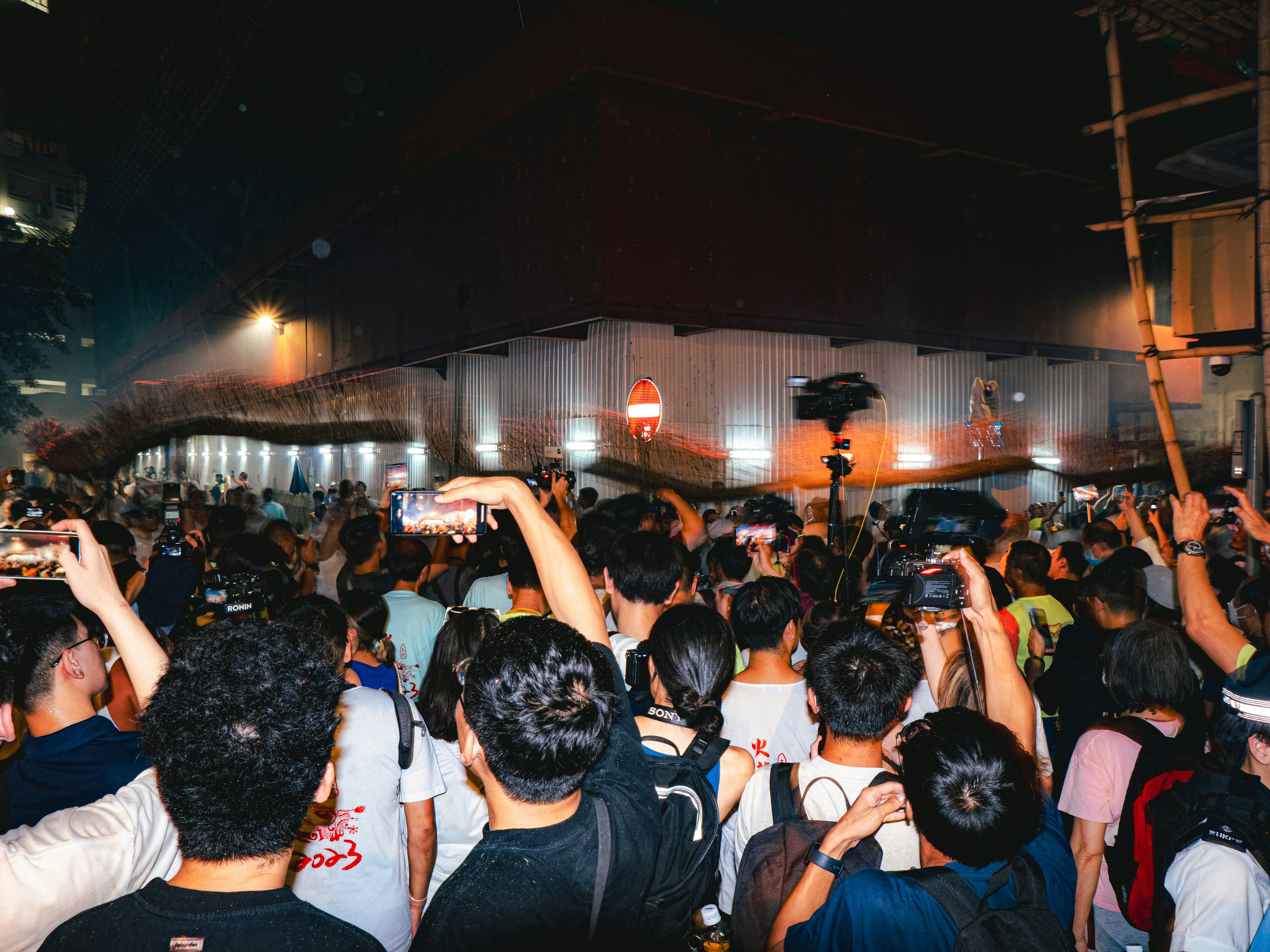 Dense night-time crowd at street event with cameras raised and a backdrop of urban lights; a concert-like, candid scene.