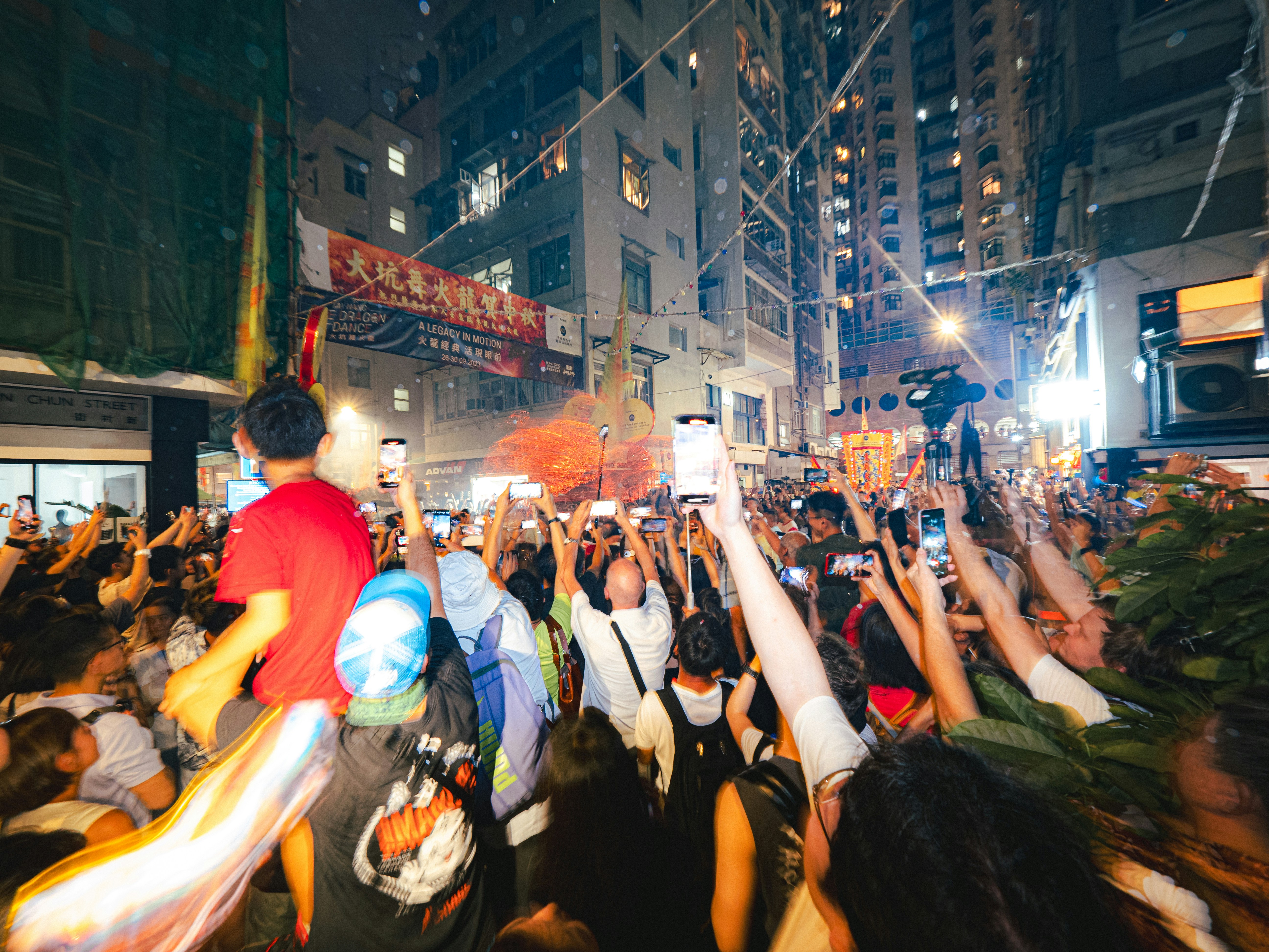 Energetic urban crowd gathered in a narrow street during a night protest, raised hands and phones capturing the moment; a banner and city lights create a vibrant backdrop. This is a photographic capture of a live event, not a painting or illustration.