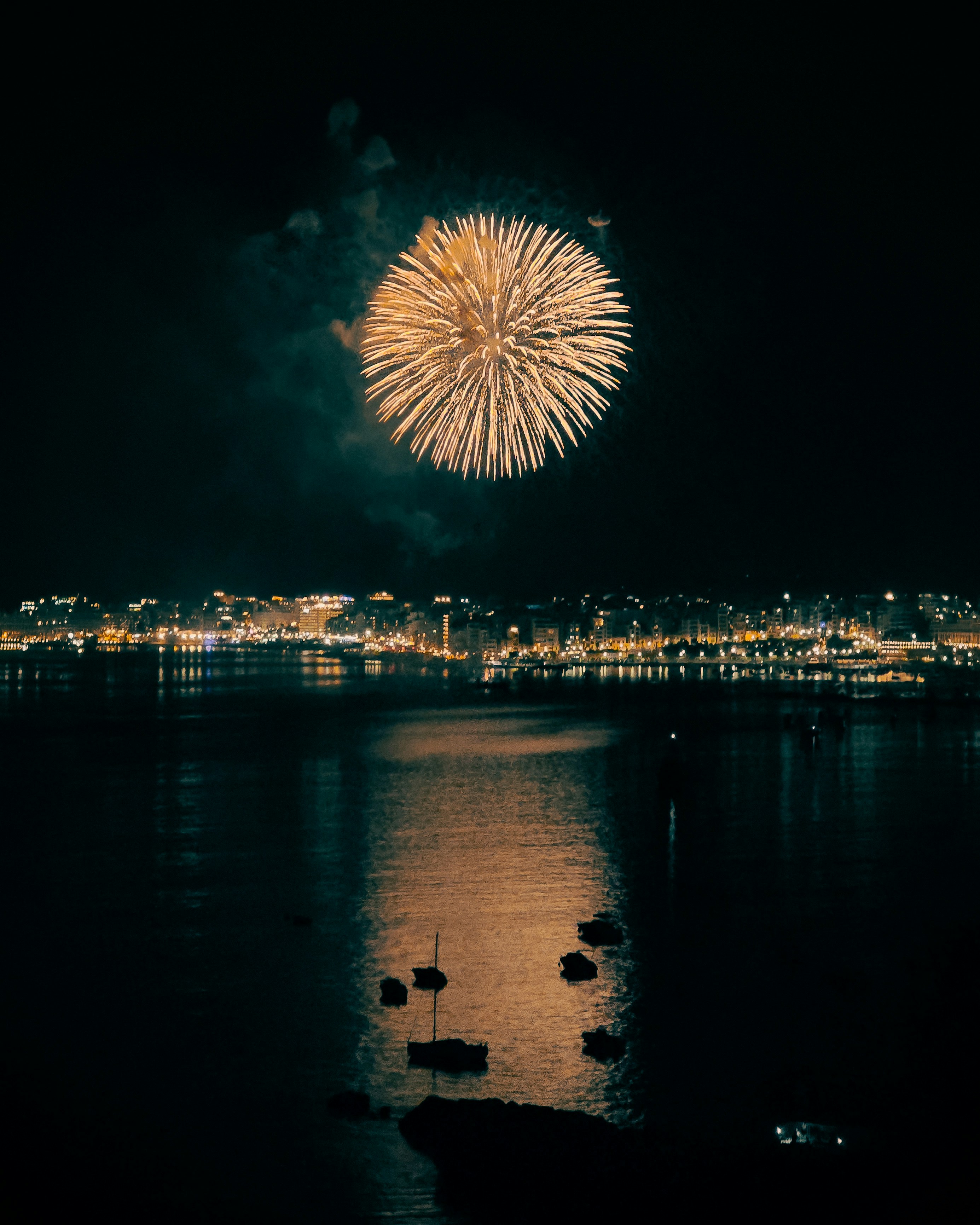 A golden firework exploding over a dark bay with boats and city lights