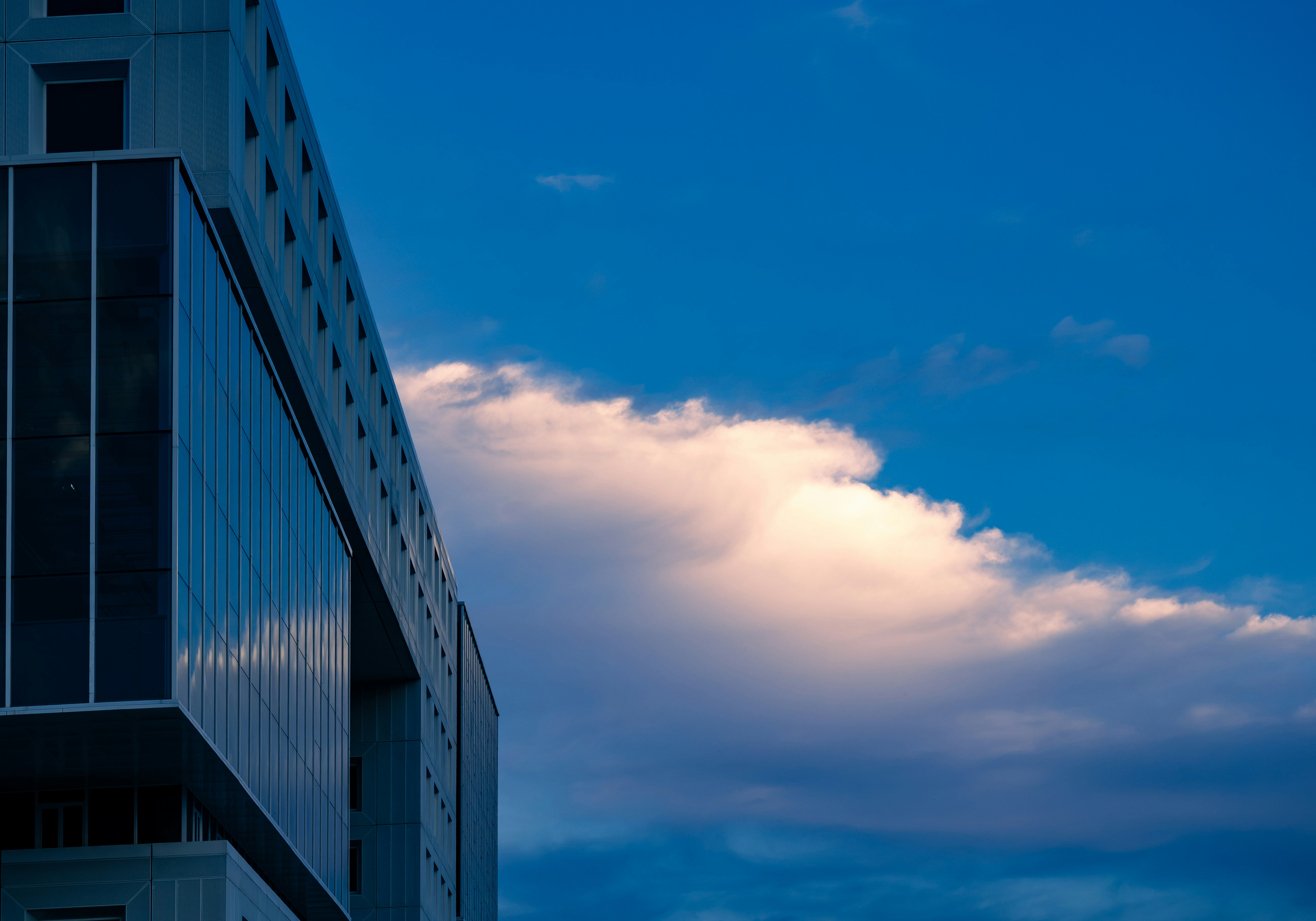 Architectural detail of a modern glass-clad building against a dramatic blue sky with a luminous cloud, emphasizing structure and mood. (JPEG logo-free stock-style photography.)