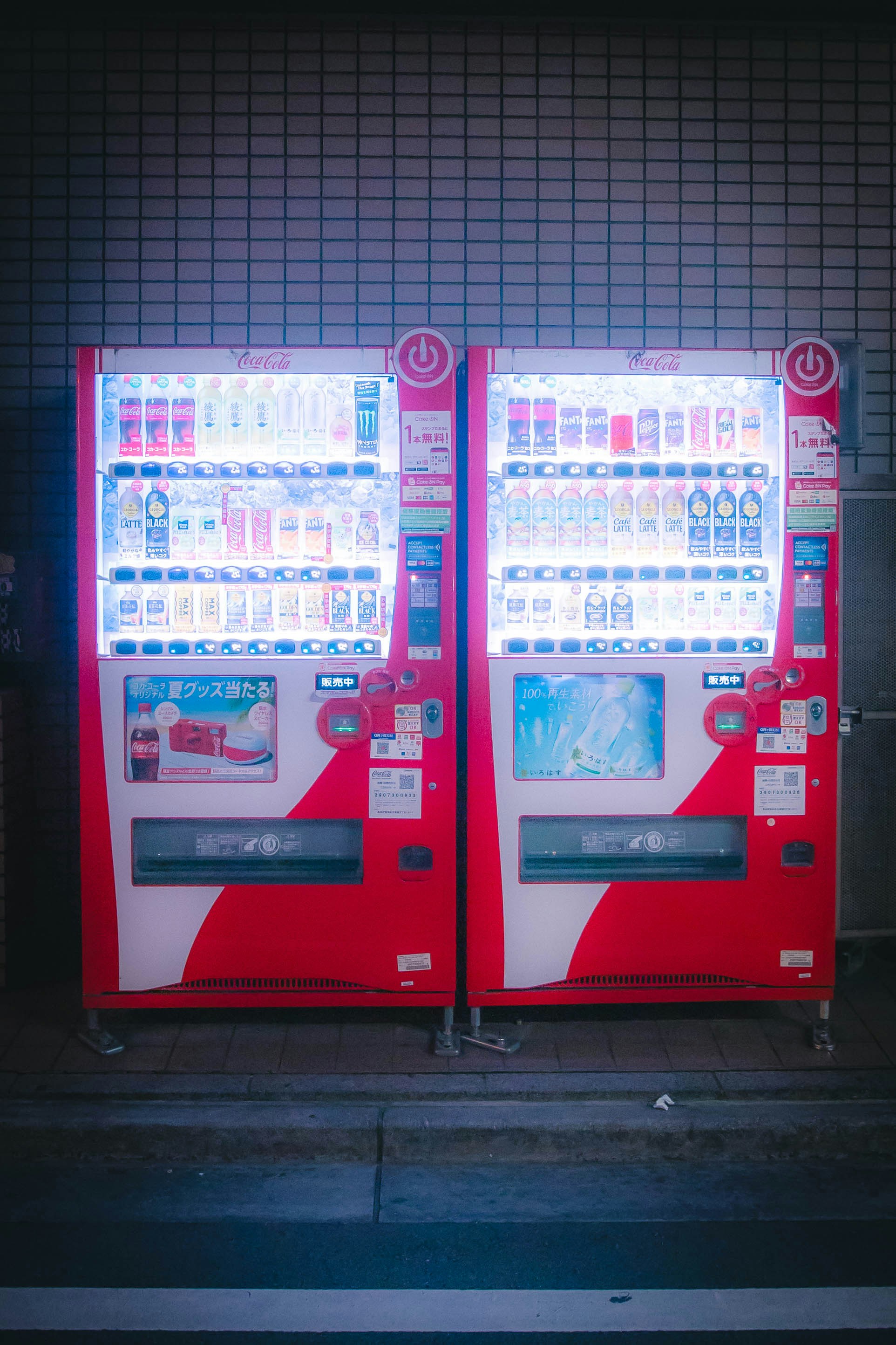 Two bright red vending machines line a dim sidewalk, their illuminated panels displaying beverages under a moody dusk glow.
