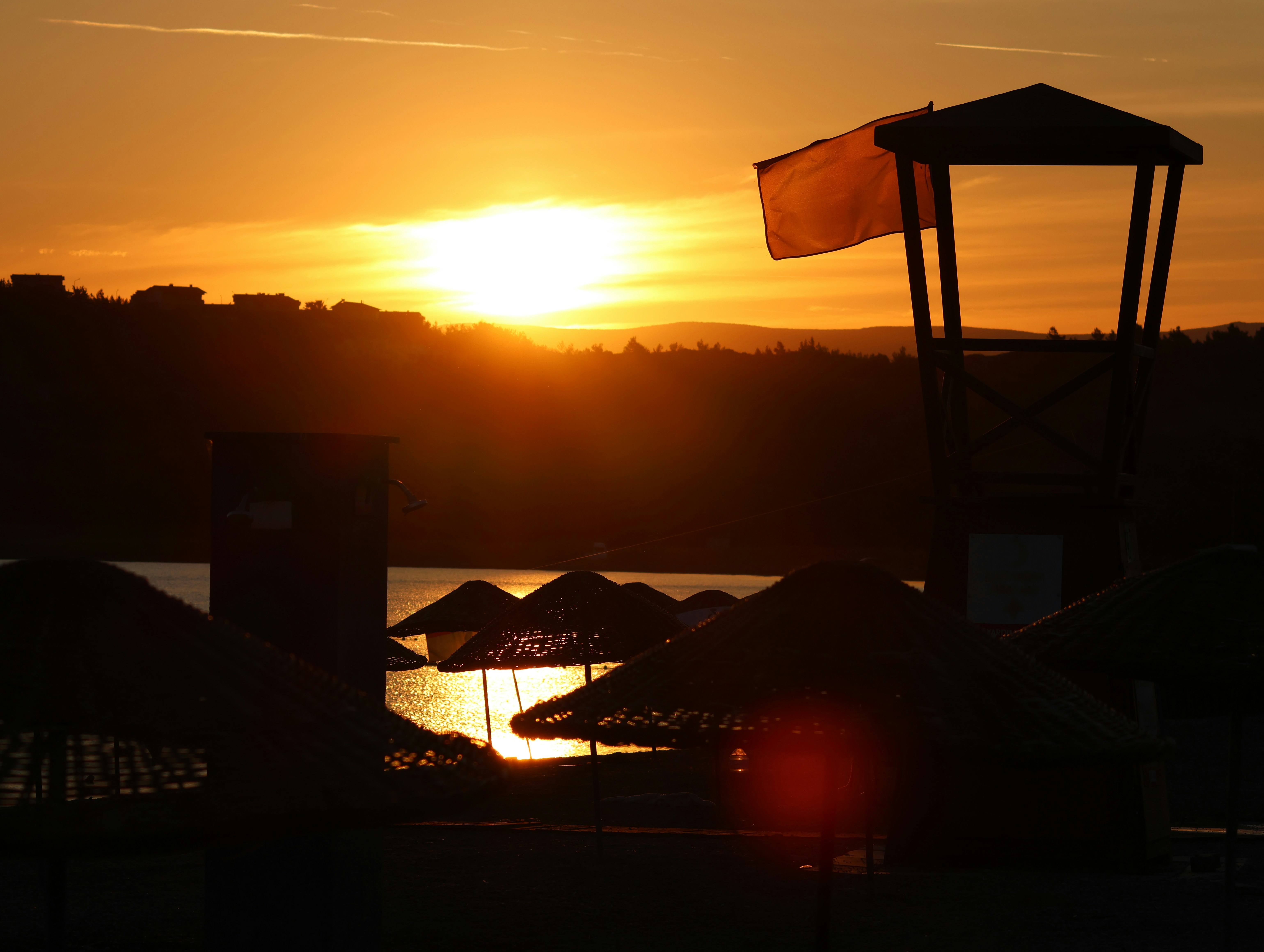 Silhouetted lifeguard tower and beach cabanas fade into a glowing sunset along the shore, with warm orange hues reflecting on calm water. A logo-like structure hints at a resort environment.