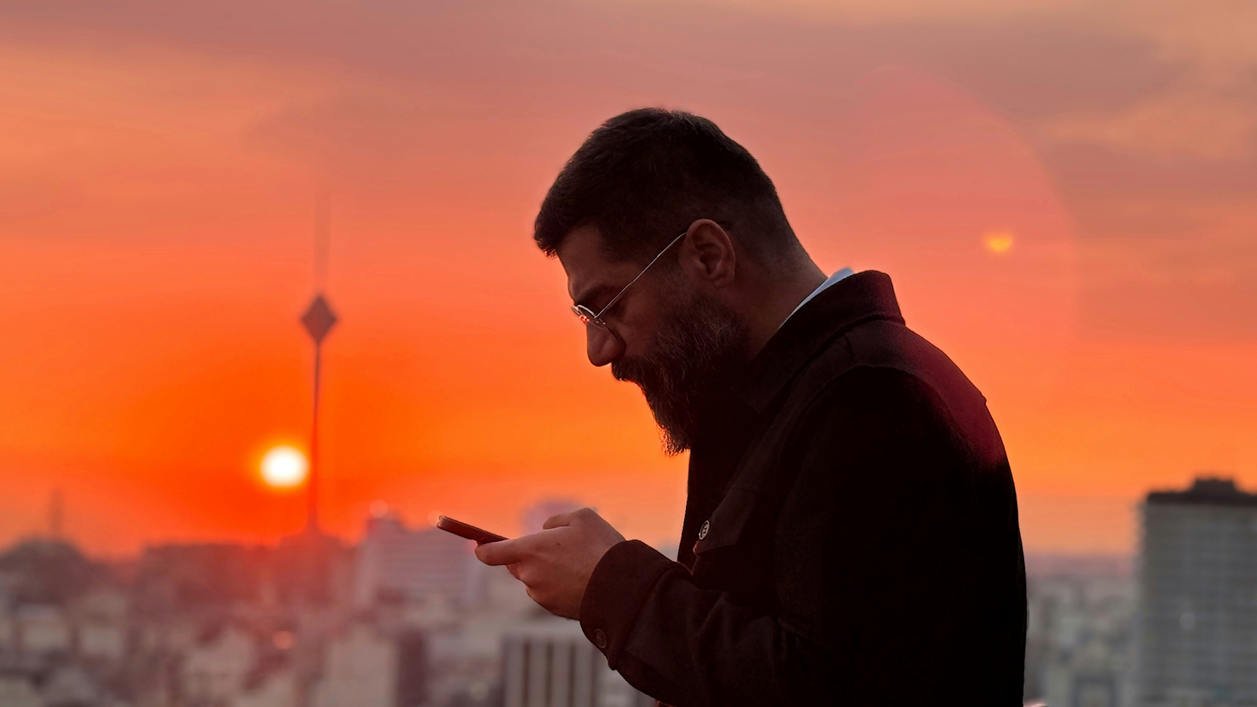 Person with glasses stands in profile at sunset, eyes down toward a phone; urban skyline and a warm orange sky provide the backdrop. Identifies a portrait-like scene with a smartphone as the focal activity.