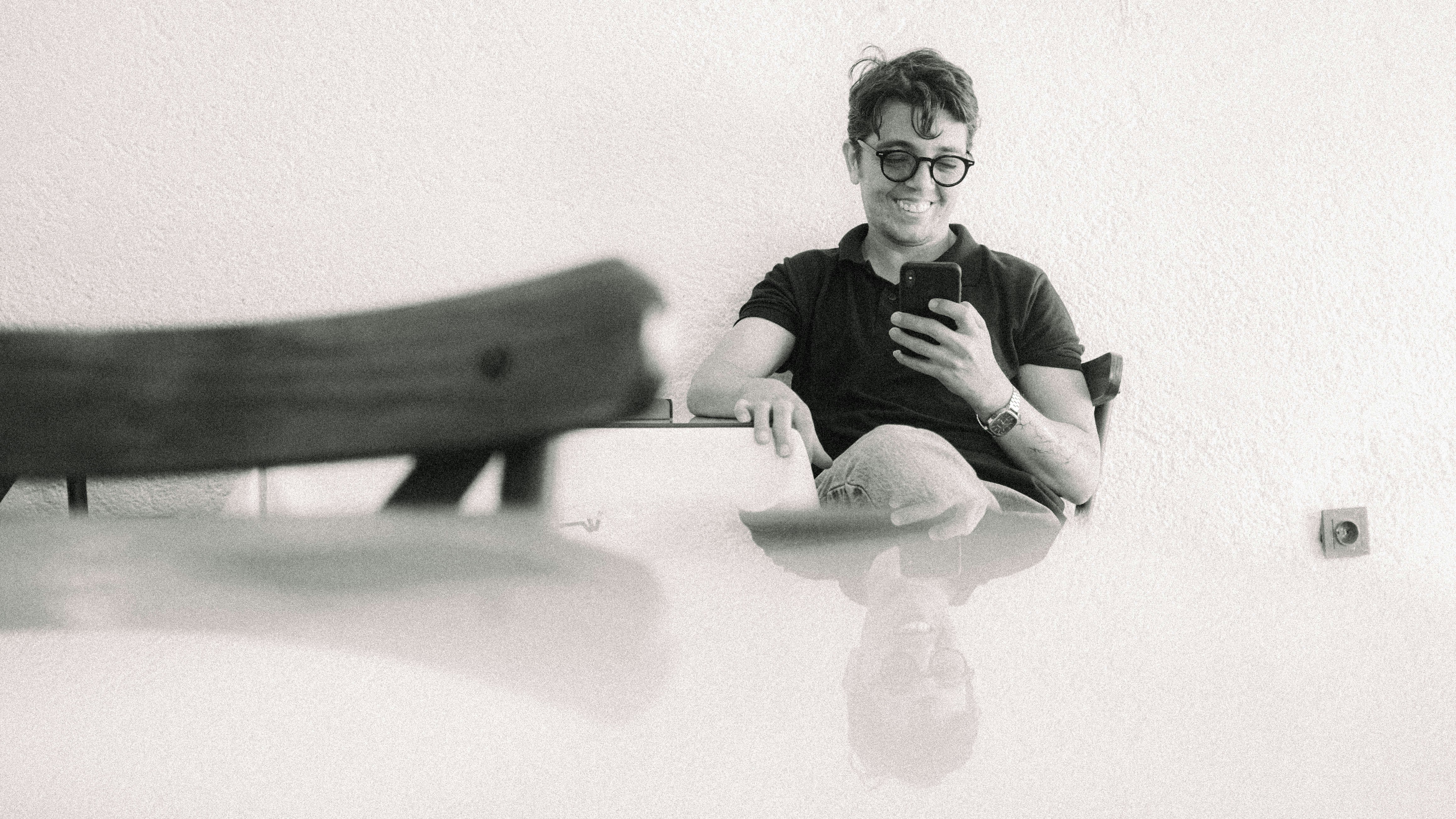A young man sits against a white wall smiling at his phone, his reflection caught in the polished floor.