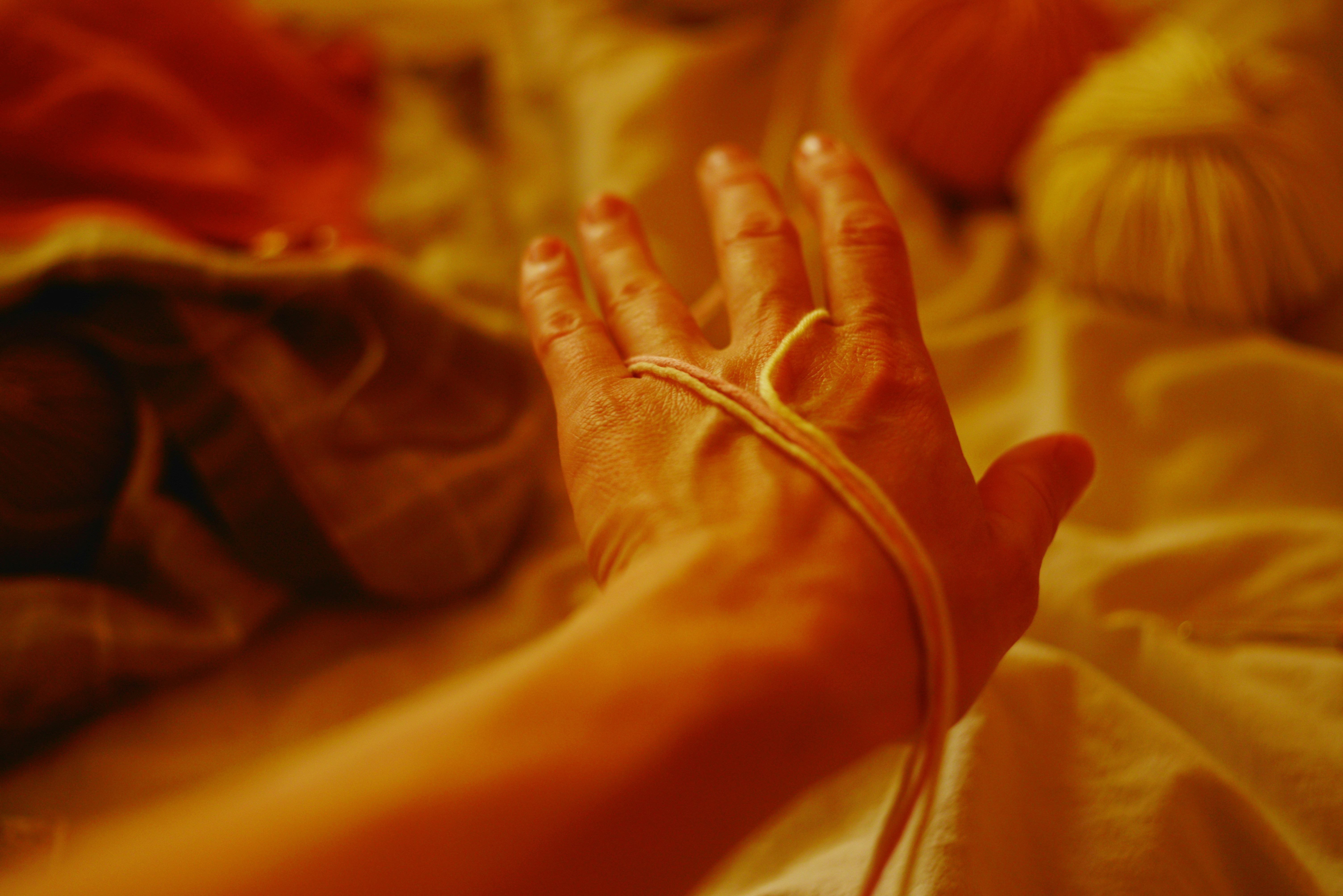 A close-up of a hand bound with a light cord against a warm, blurred bed-linen backdrop, suggesting restraint and intimacy; the scene centers on the fingers and rope texture. This is a staged, intimate still rather than a candid moment.