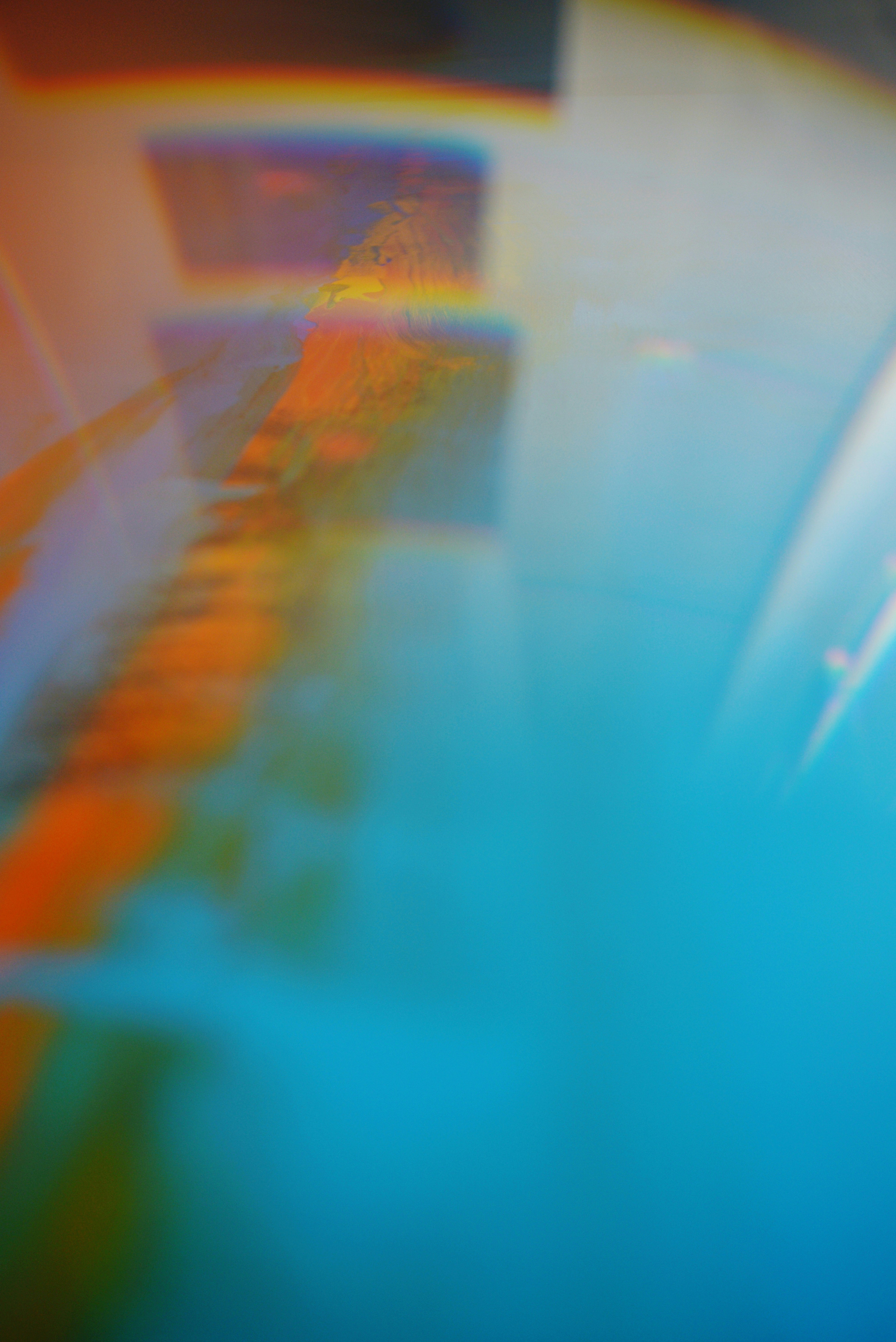 Softly blurred, multicolored light streaks fold into a dreamy gradient; a painterly mix of blue, orange, and green creates an impressionistic abstraction with no discernible subject. This appears to be a diffused reflection or refraction scene, not a concrete object.