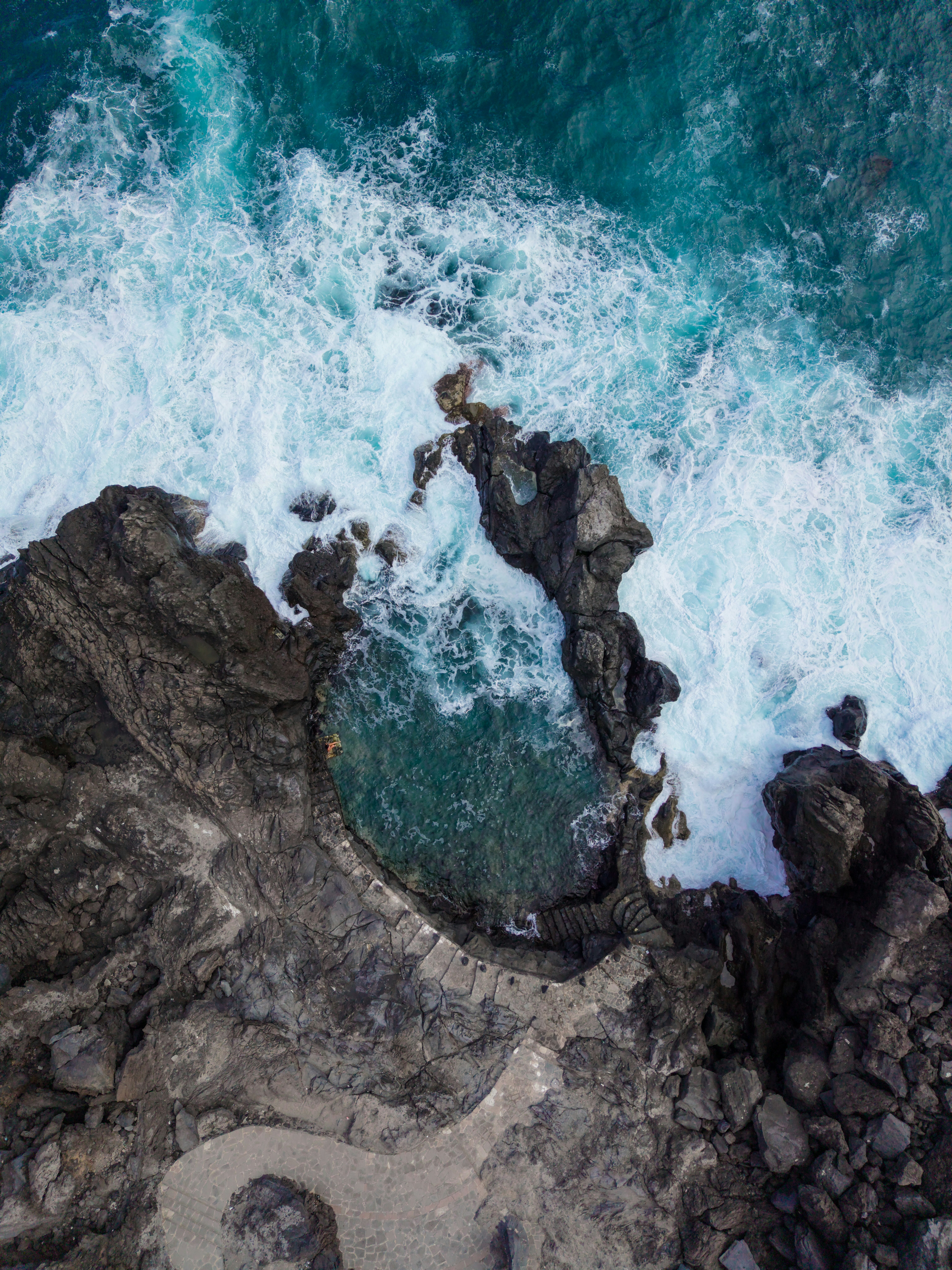 Aerial shot of dark volcanic rocks meeting turquoise surf; foamy white water swirls into a natural inlet creating a dramatic coastal formation.
