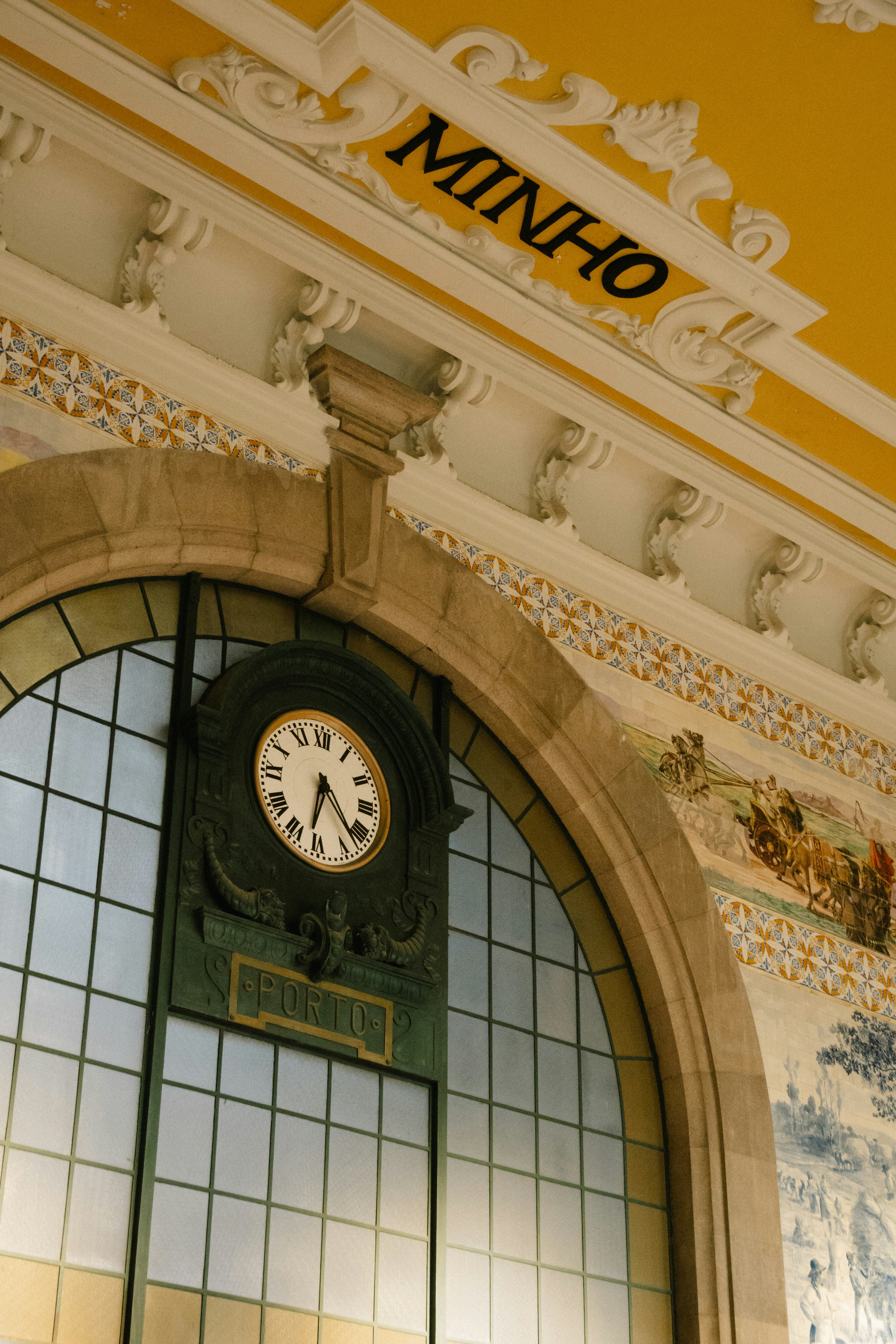Arched windowed clock facade under ornate detailing and a yellow sign reading 'MINHO' in a historic station interior. A decorative scene with tile patterns and framed mural to the right.