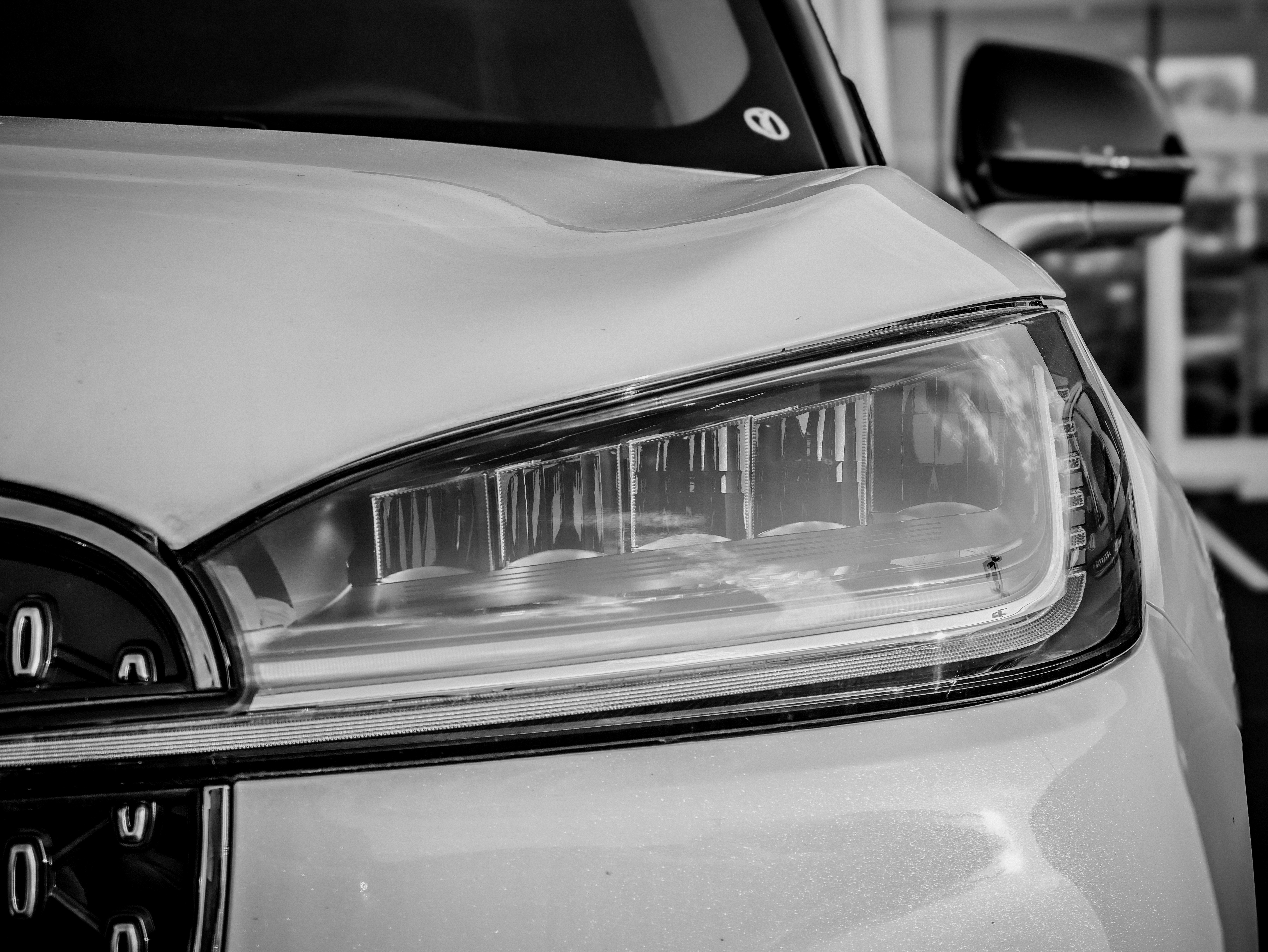 This crisp monochrome macro photograph highlights an intricate automotive headlight assembly. Four distinct square LED projector lenses rest behind polished glass, contrasting cleanly against the smooth sweeping metal hood panel.