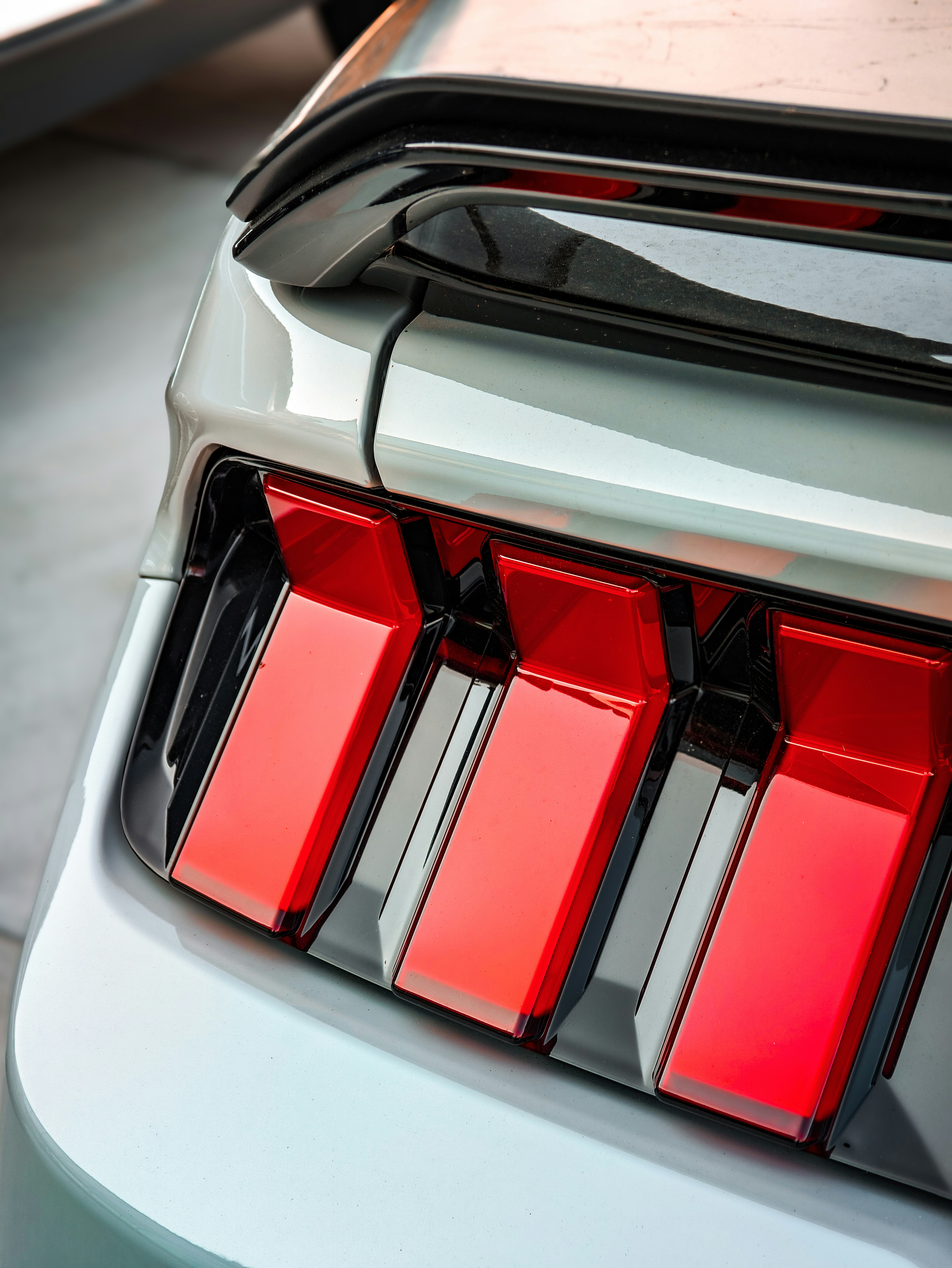 This detailed macro photograph captures a modern grey sports vehicle featuring three distinct vibrant red vertical taillight bars set deeply within a highly polished glossy black panel under a spoiler.