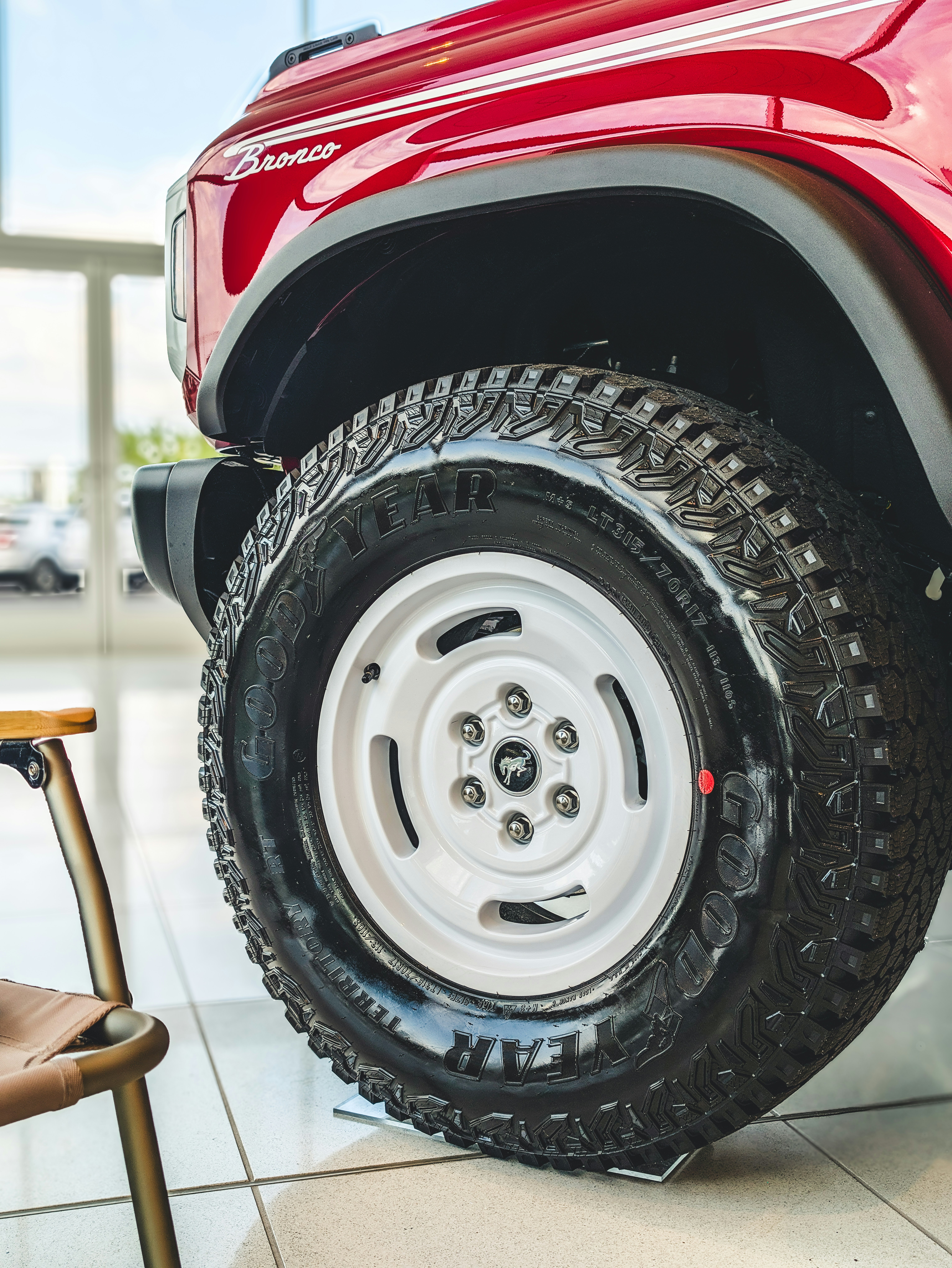 A glossy red Bronco fender with crisp white retro pinstriping arches over a rugged Goodyear tire mounted on a classic solid white alloy wheel upon a bright tiled showroom floor.