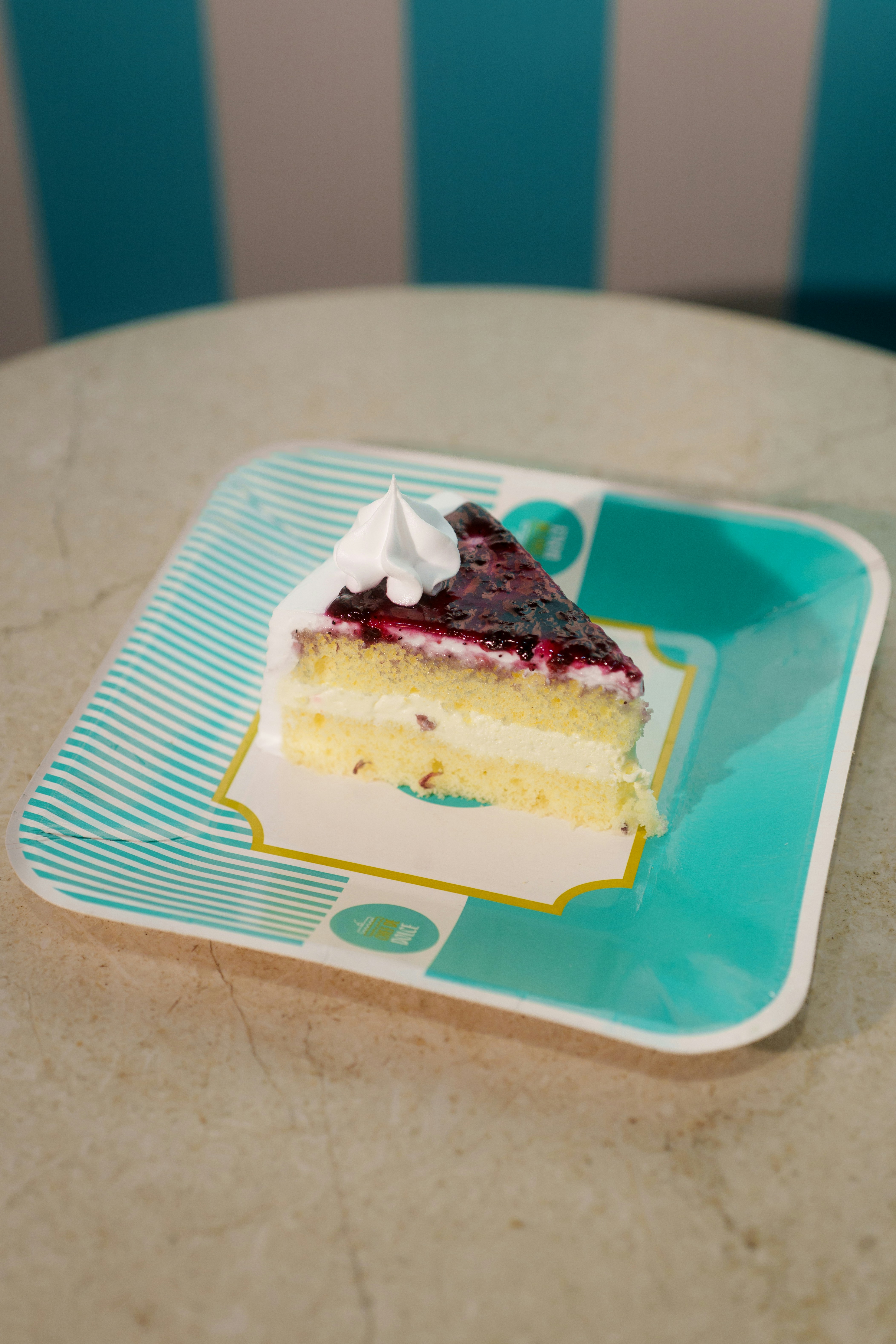 Dessert slice with whipped cream sits on a turquoise patterned tray, showcasing layered cake and berry topping on a rounded plate.