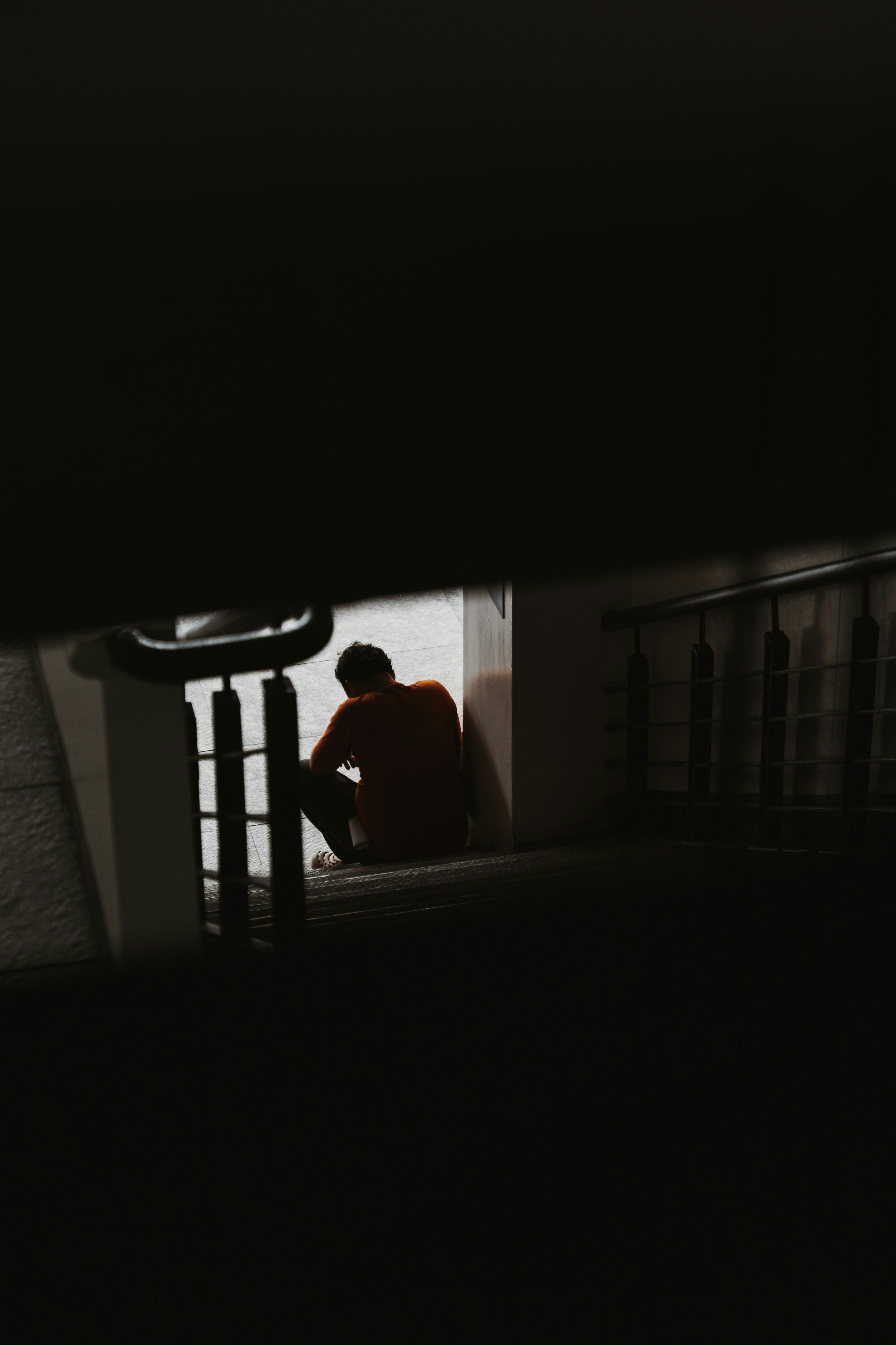 A lone figure sits in a dim stairwell doorway, backlit by a bright exterior, creating a moody, contemplative scene with strong contrast and a sense of isolation.