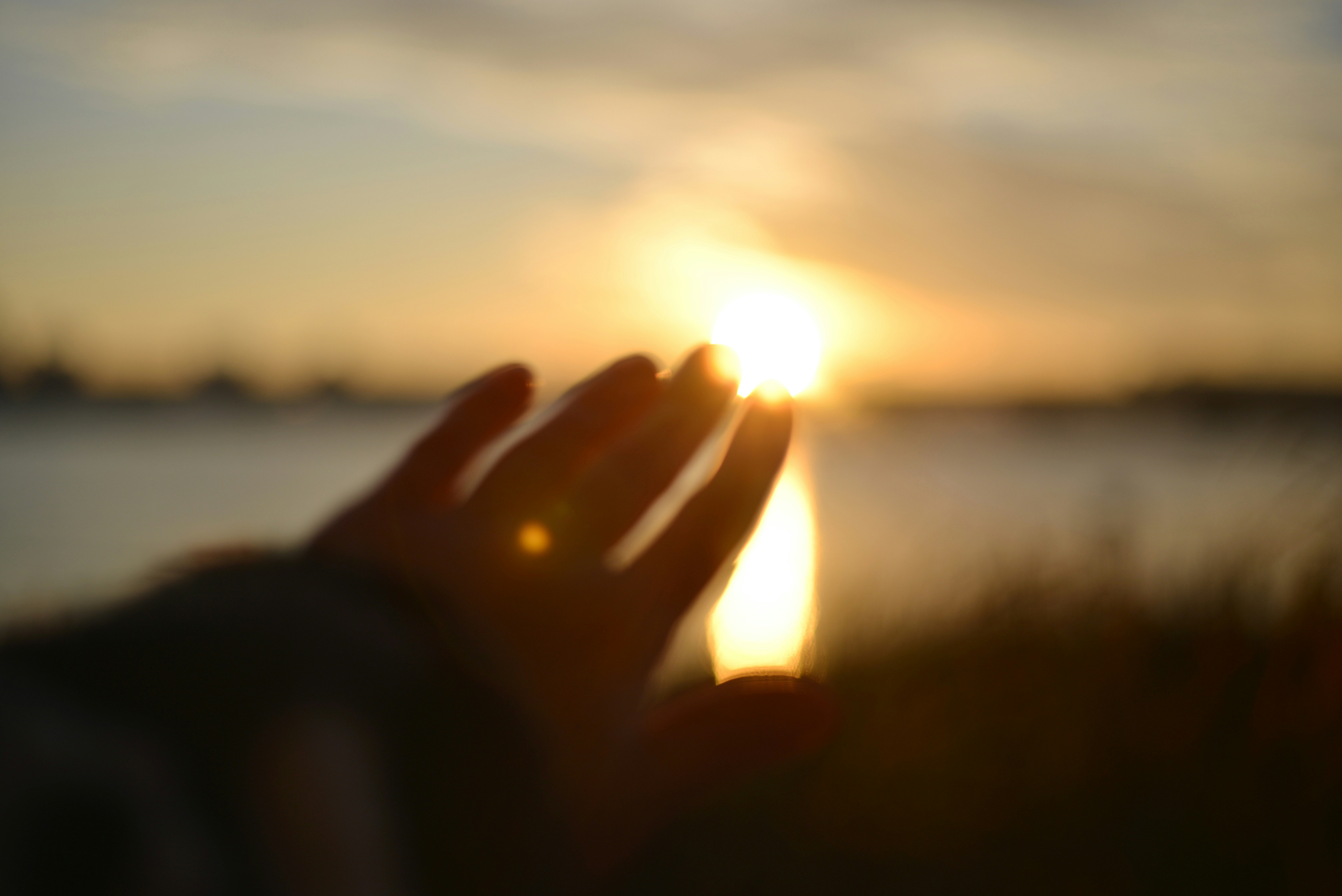 Blurry foreground hand reaching toward a low sun over a calm body of water; warm backlight and soft bokeh create a dreamlike silhouette, suggesting a moment of connection or release. This is a photographic capture, not a graphic or illustrator.