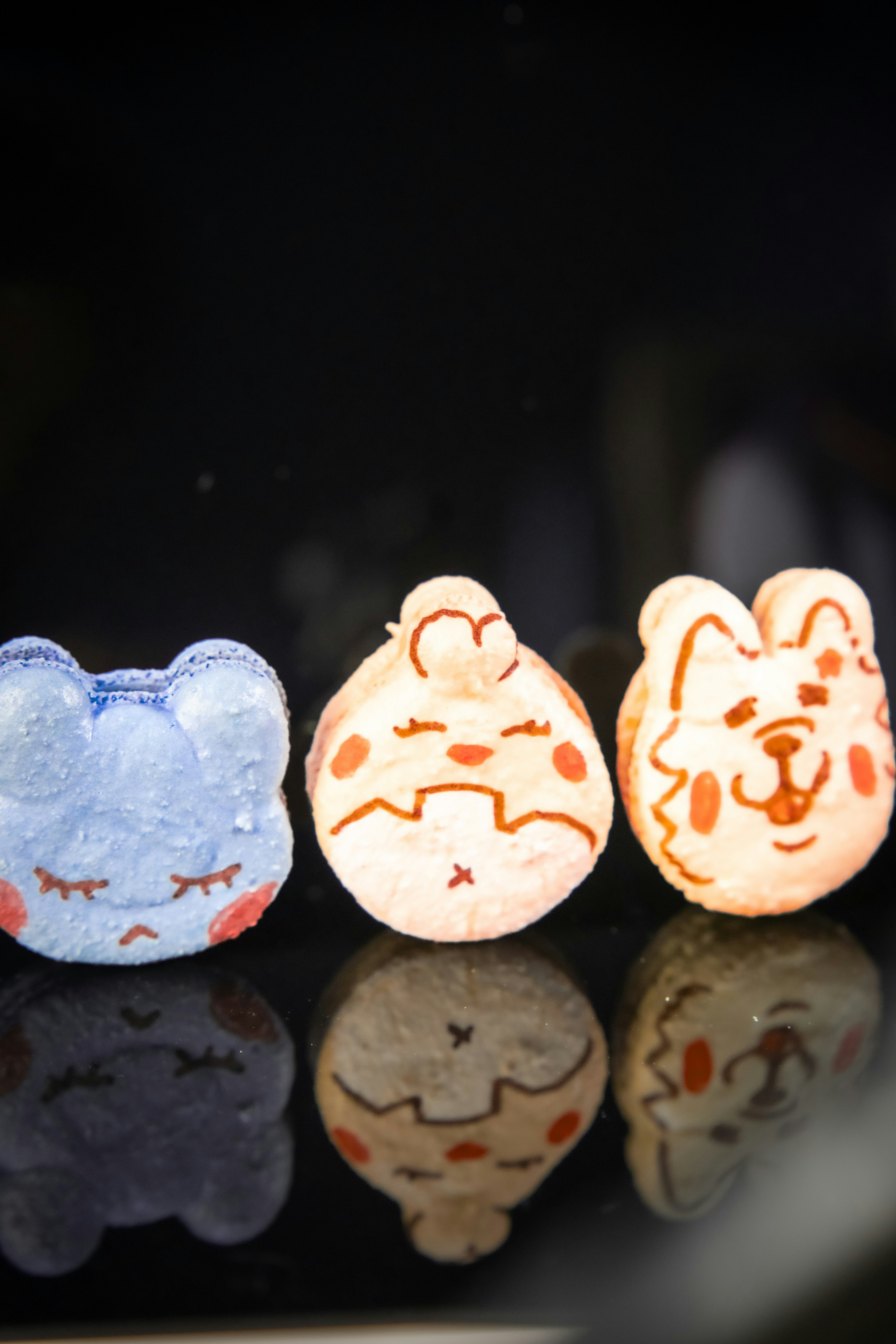 Three animal-shaped macarons with painted faces on a reflective black surface