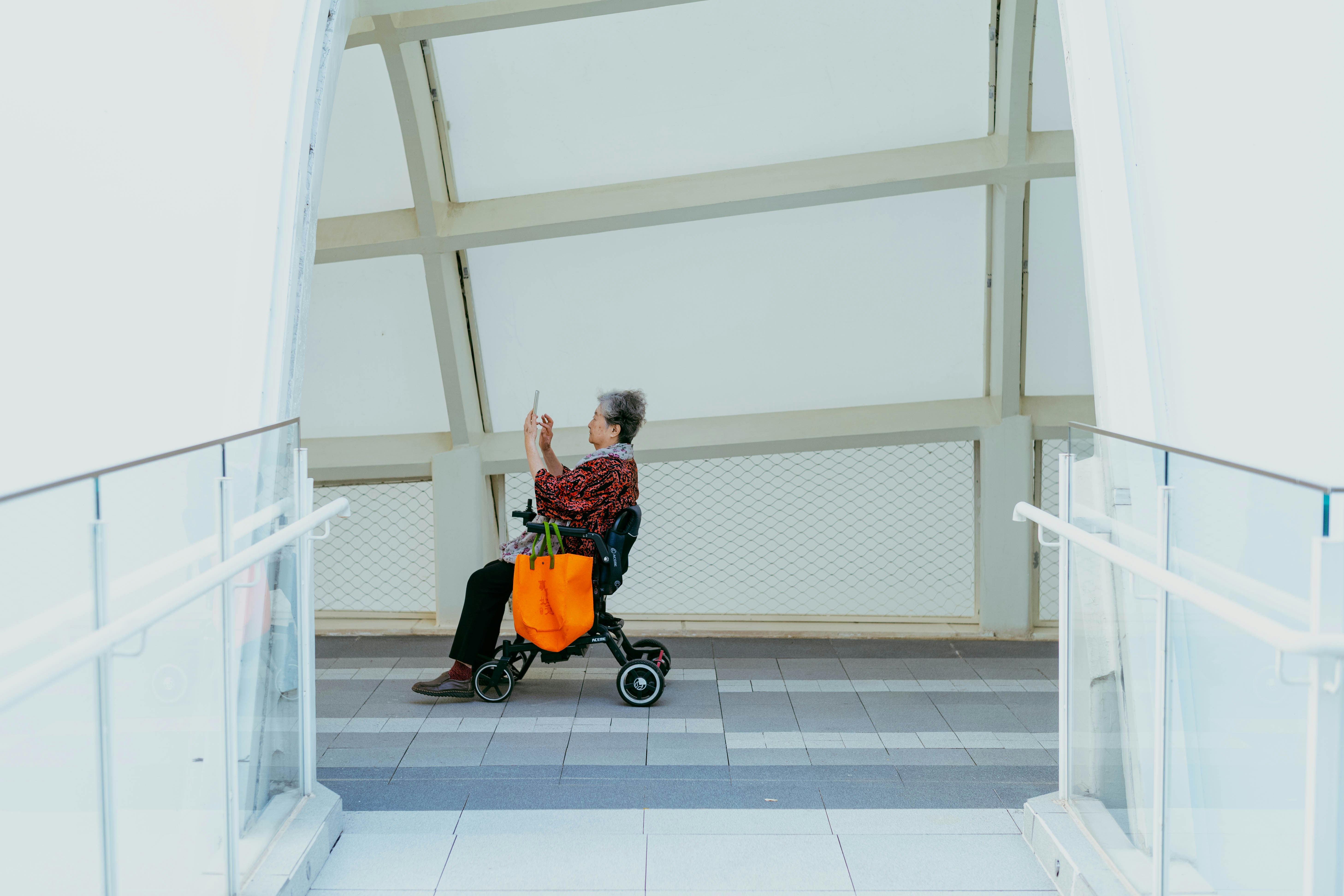 A person in a wheelchair sits in a bright, minimal atrium, gesturing with one hand raised, orange bag resting beside; clean lines and soft light emphasize calm, open space.