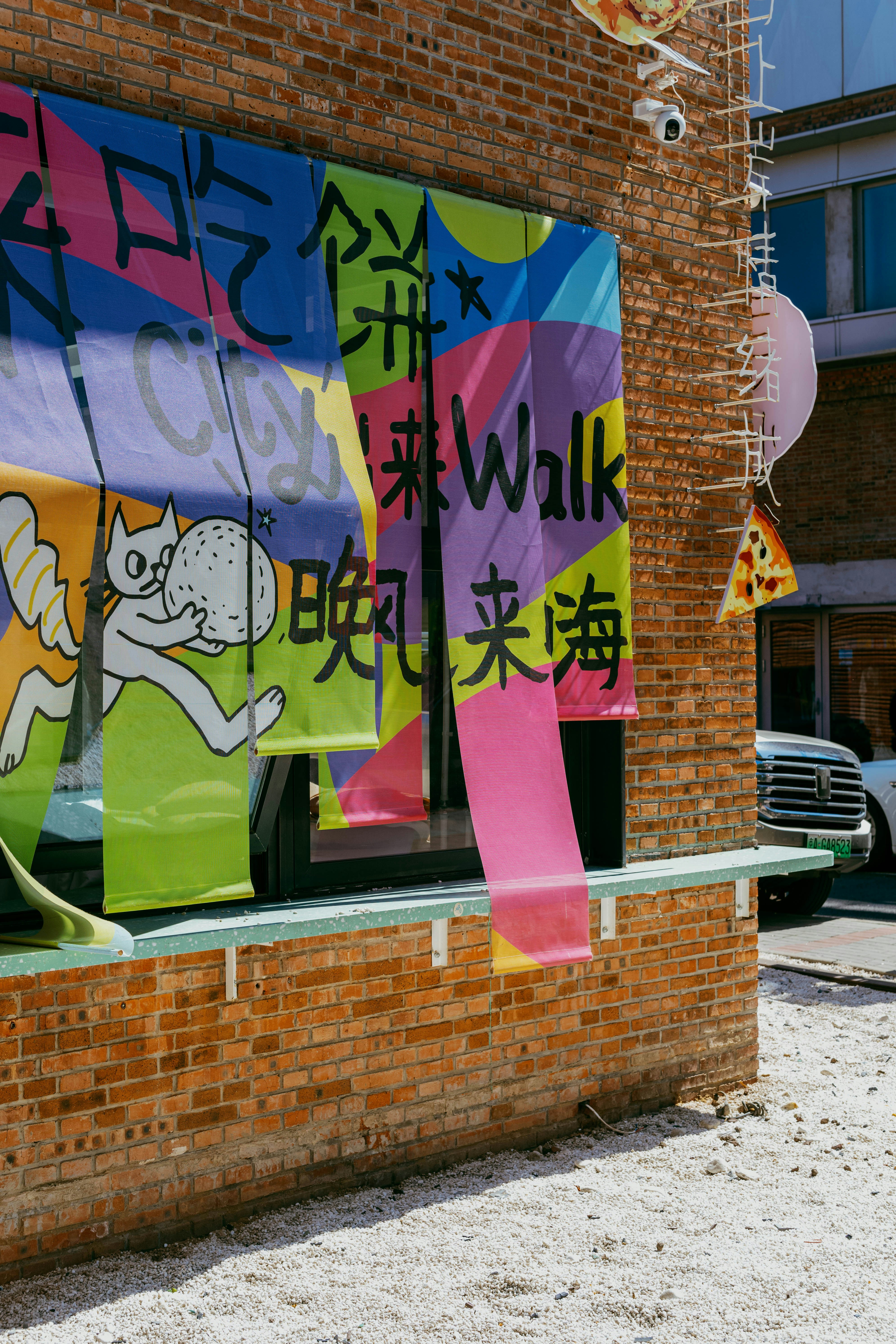 Bright mural panels wrap a brick storefront, featuring a cartoon cat and bold Chinese characters; sunlight highlights the painted glass and a pink ribbon draped out the window.