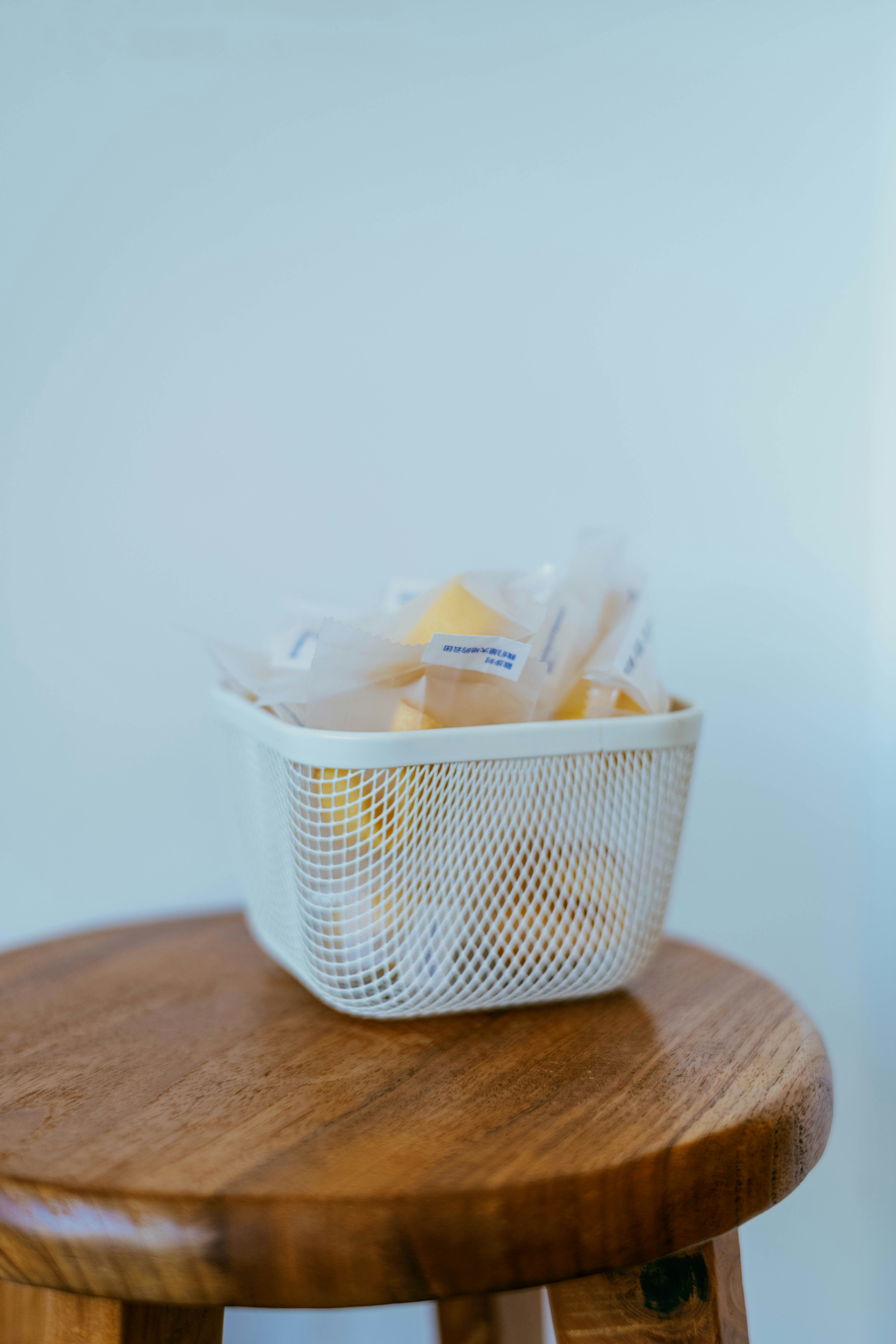 A white mesh basket filled with small packets sits atop a round wooden stool against a soft, blurred background. This is a still-life photography setup featuring a simple domestic object.
