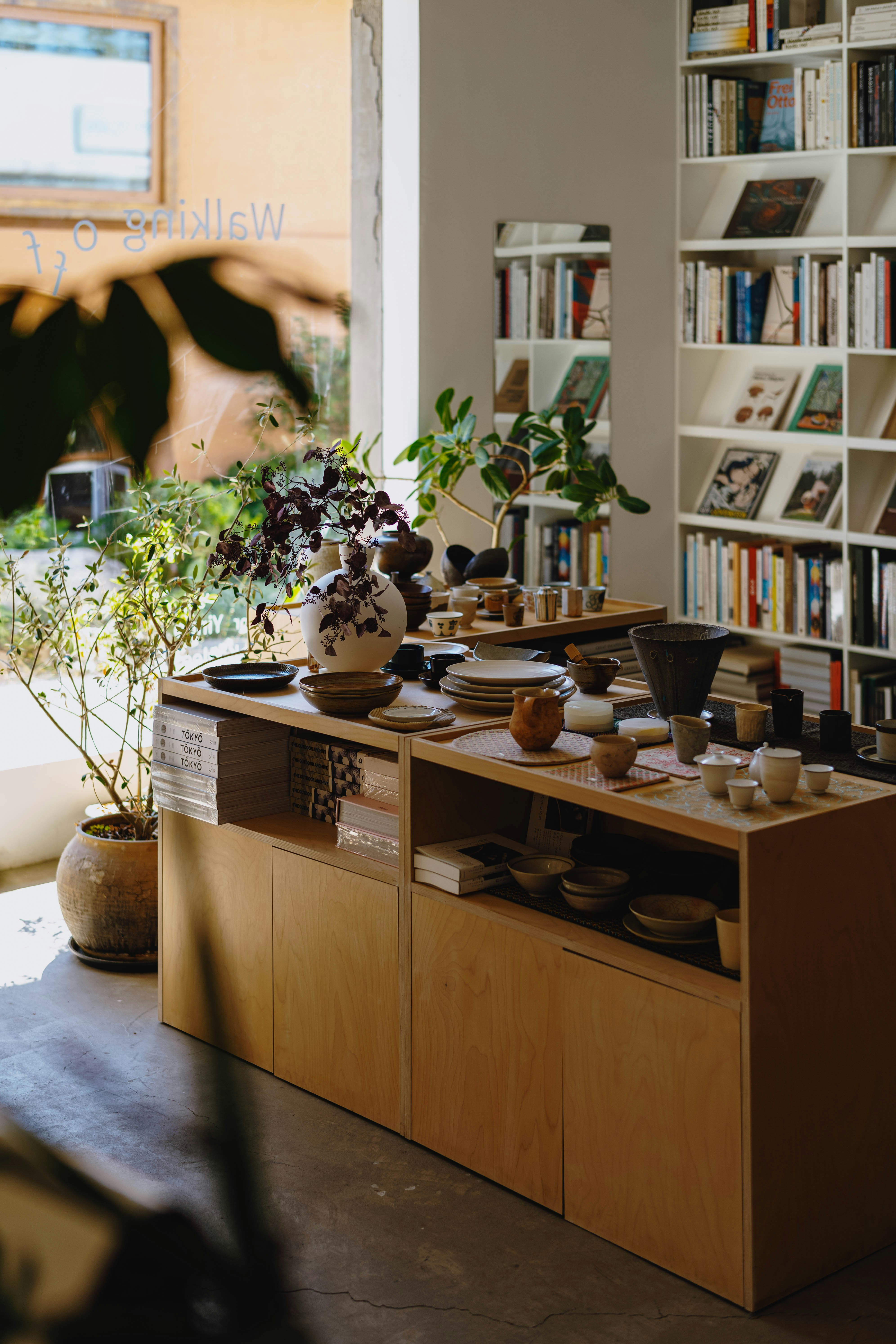 Warm still-life scene inside a bookshop-like space, featuring a wooden counter with ceramic bowls and plants, backed by a wall of shelves filled with books. A calm, curated display emphasizing craft and texture.