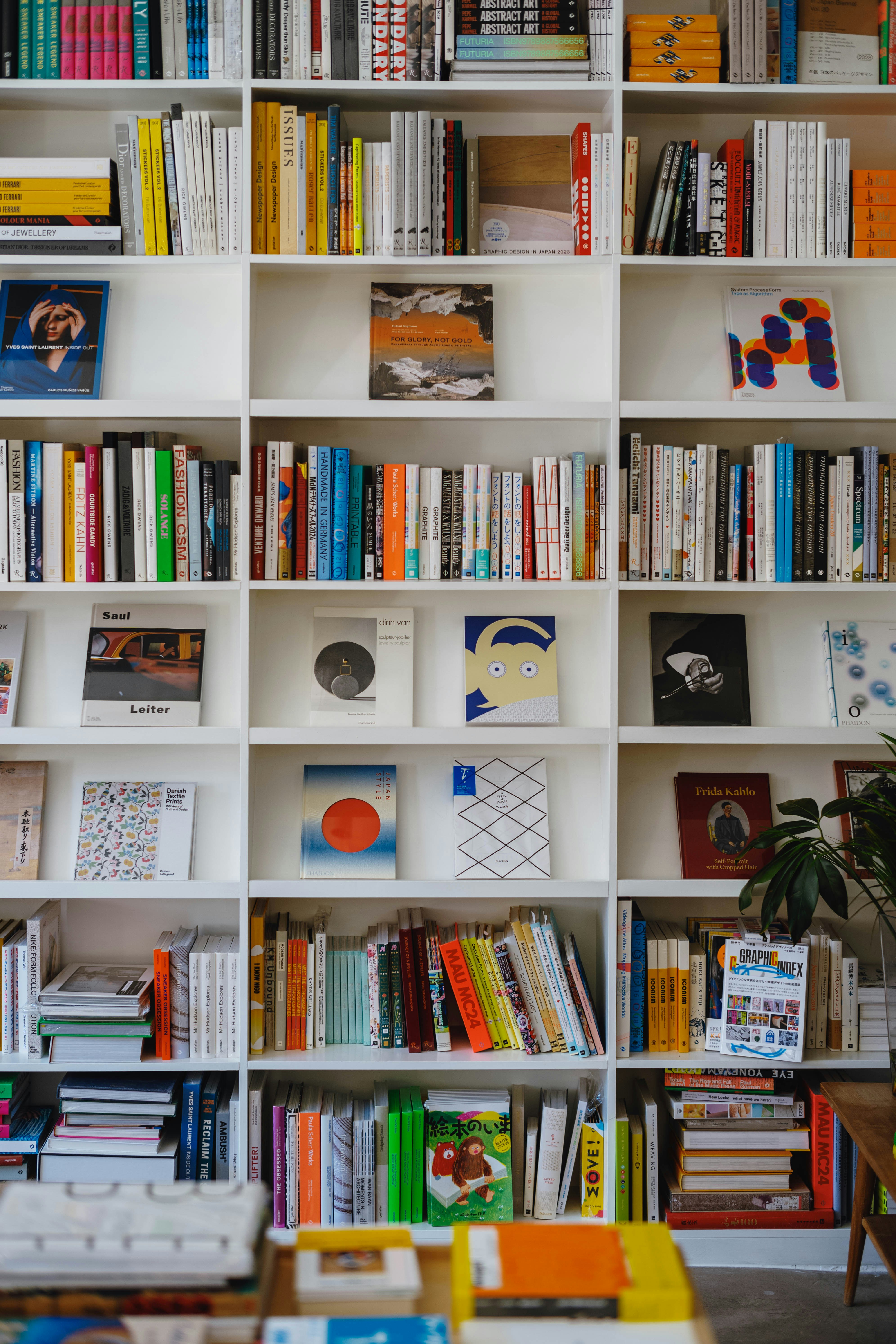 Colorful books arranged in a grid library shelf with a few art prints and plants visible, creating a calm, structured composition.