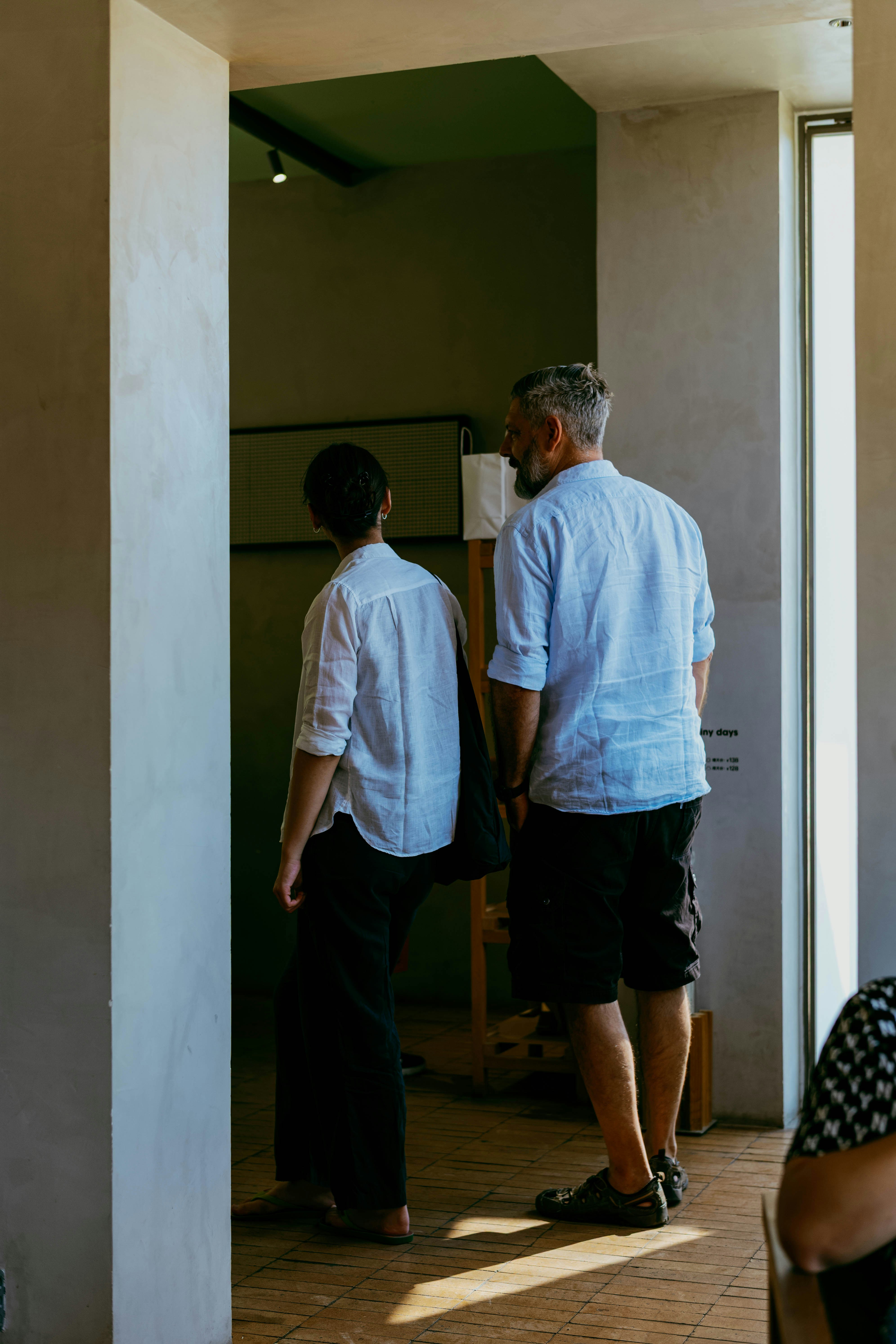 Two men in light blue shirts stand with their backs to the camera in a sunlit interior, one taller with a backpack and rolled-up sleeves. The scene focuses on their positioning near a doorway and a windowed edge, hinting at a moment of pause or transition. (This is a photograph.)