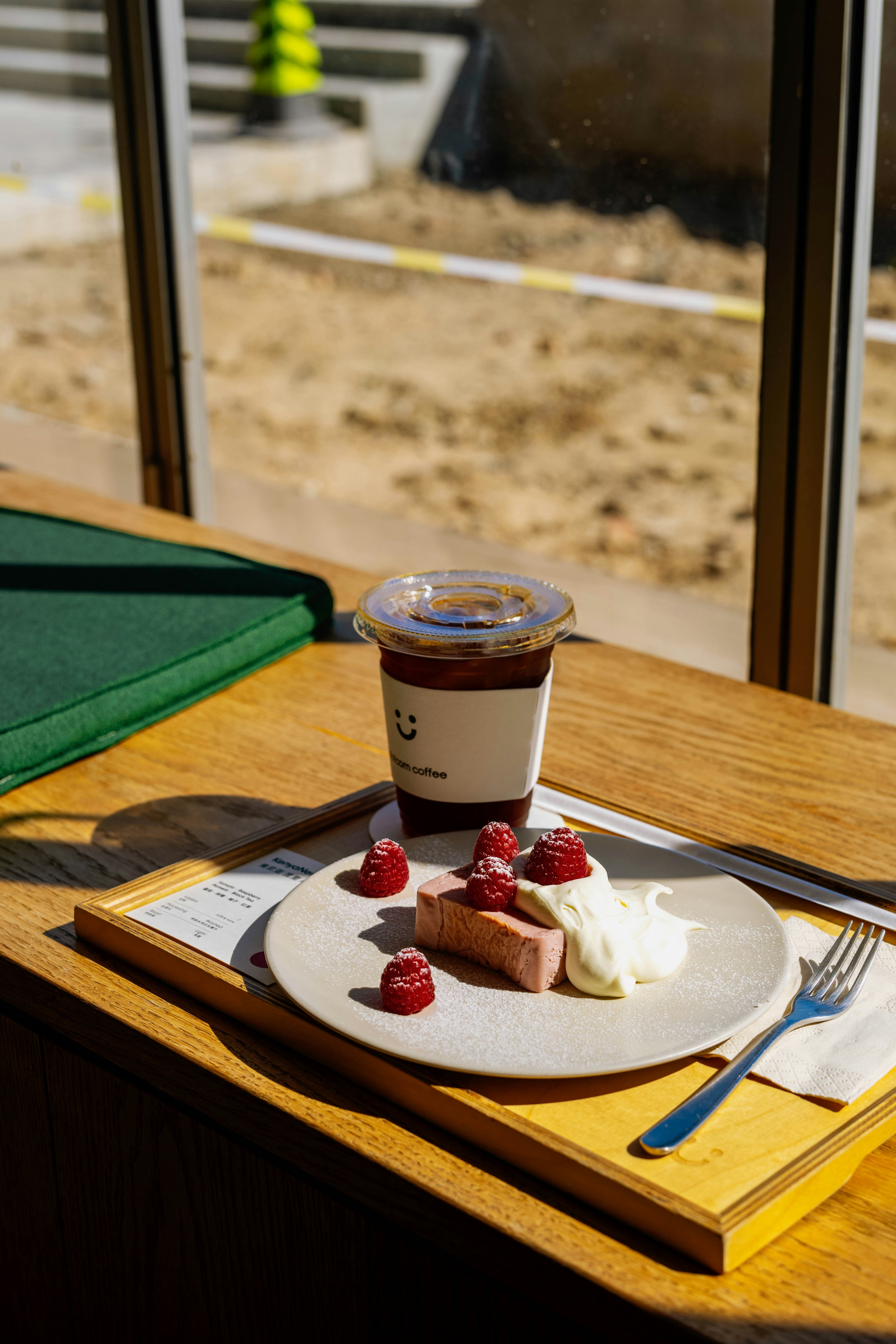Cocoa dessert plate with raspberries and whipped cream beside a takeaway coffee sits on a wooden tray by a sunlit window.
