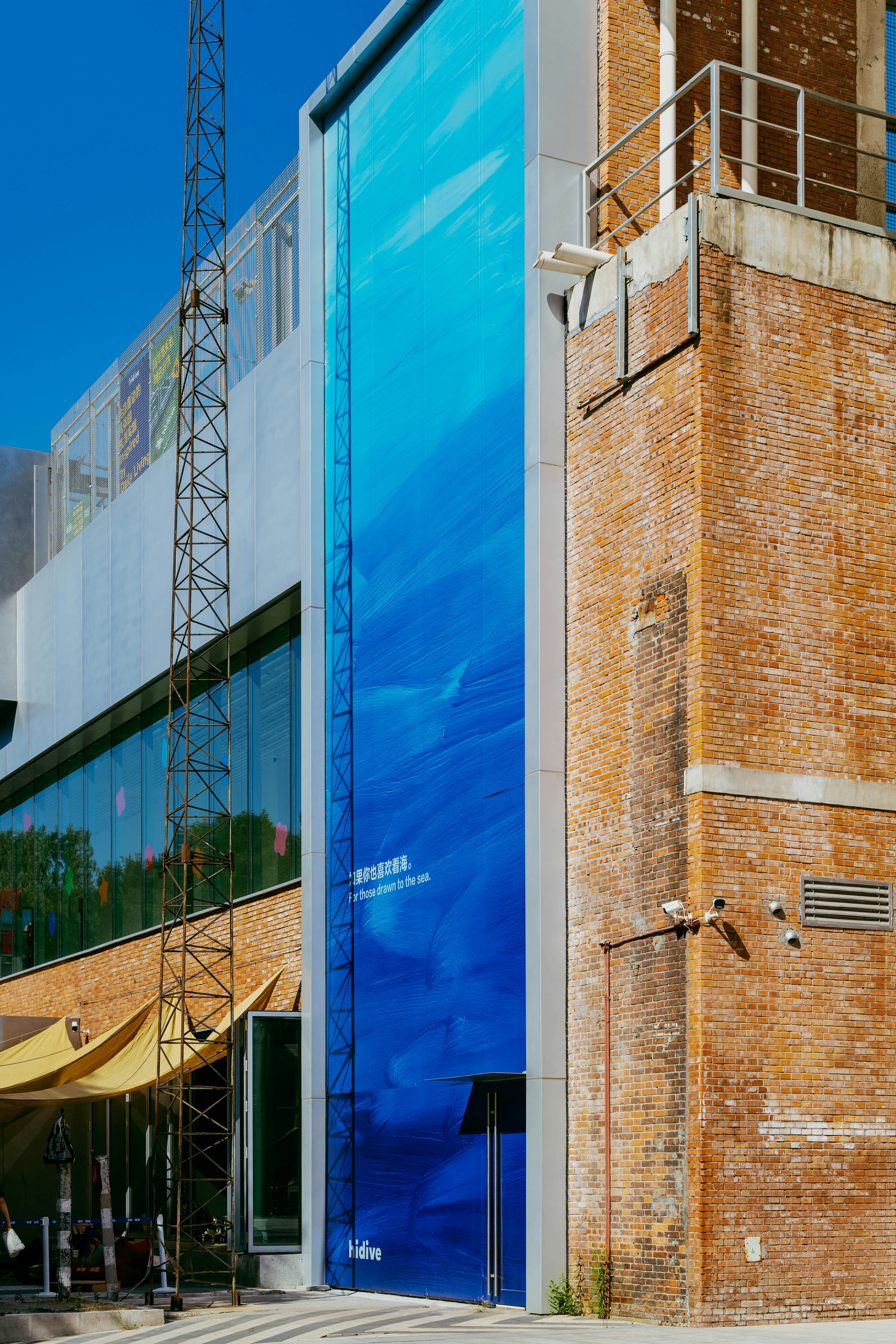 Blue-glass facade flanks a brick building, with a slender tower structure and outdoor seating beneath a sunlit canopy.