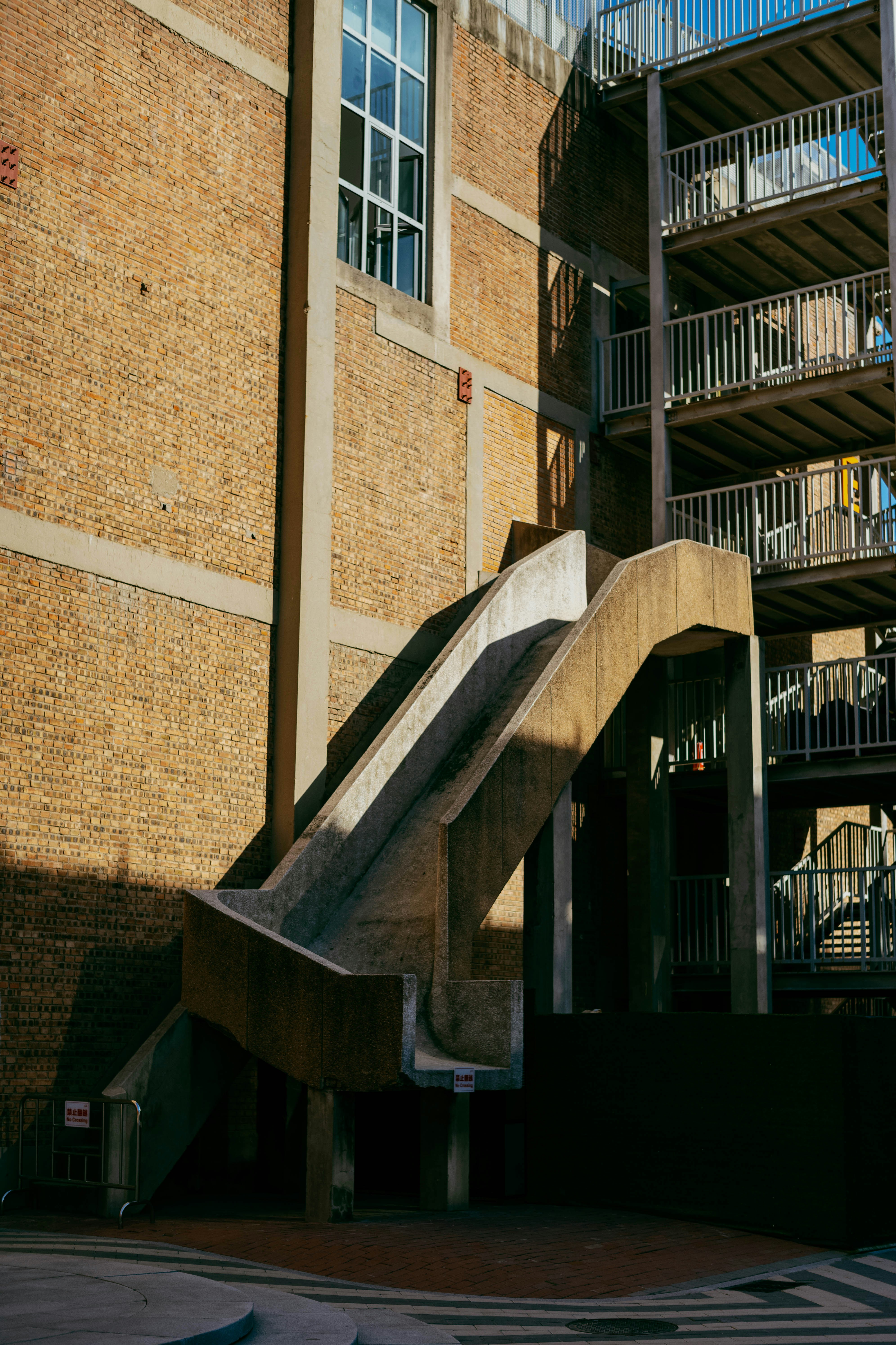 Concrete exterior staircase winding along a brick building with shadowed lower levels and metal fire escapes on the right.