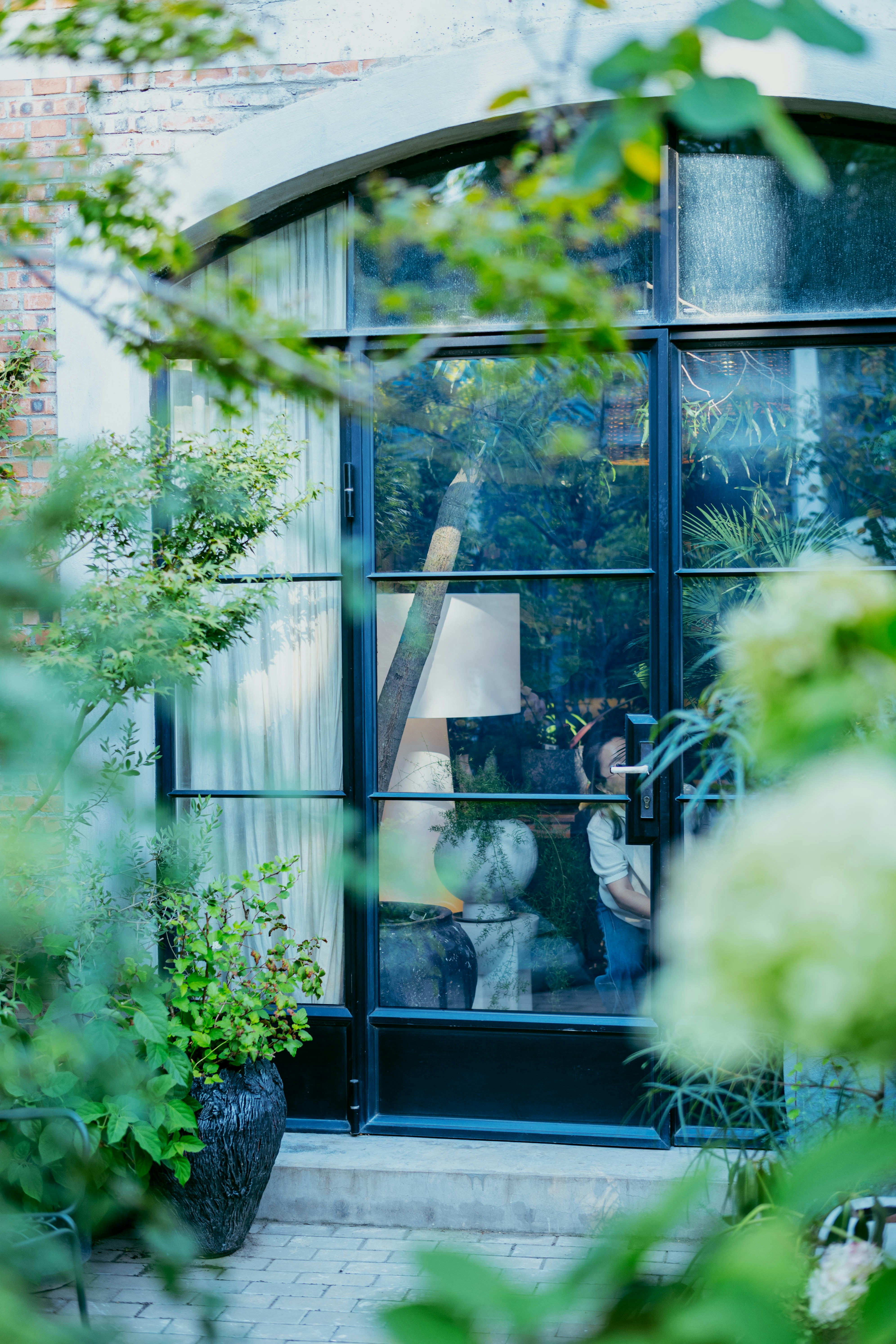 Sunlit glass doors framed by greenery reveal an interior with plants and a person inside, suggesting a tranquil garden retreat.