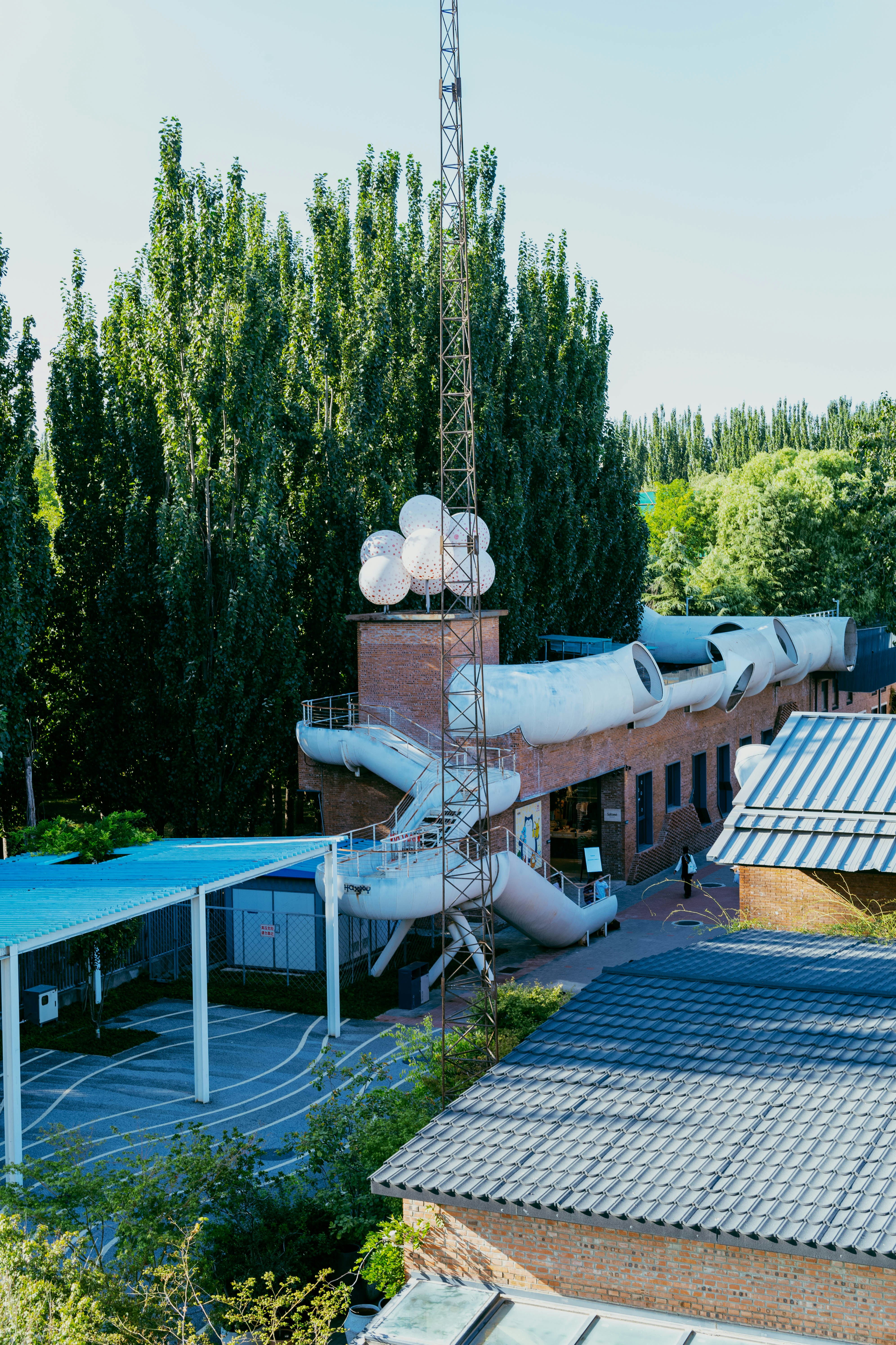 Industrial rooftop with a tall metal tower and white ductwork snaking along brick buildings; lush trees in the background indicate an elevated campus or facility. The image captures technical infrastructure and a hint of outdoor space.