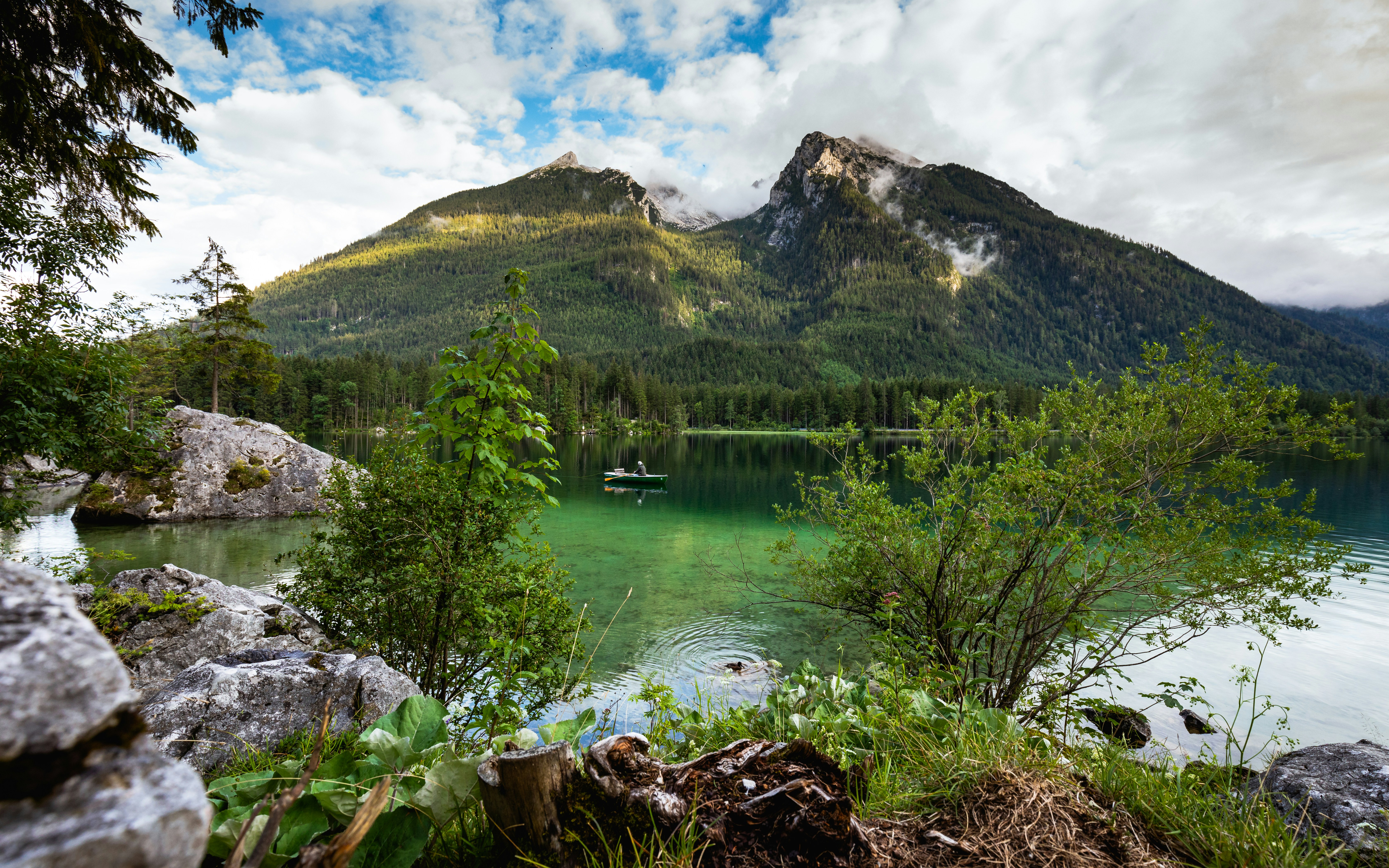 Emerald-green lake framed by rocky shore and evergreen trees with mountains in the background; a small boat rests near the shore, under a partly cloudy sky.