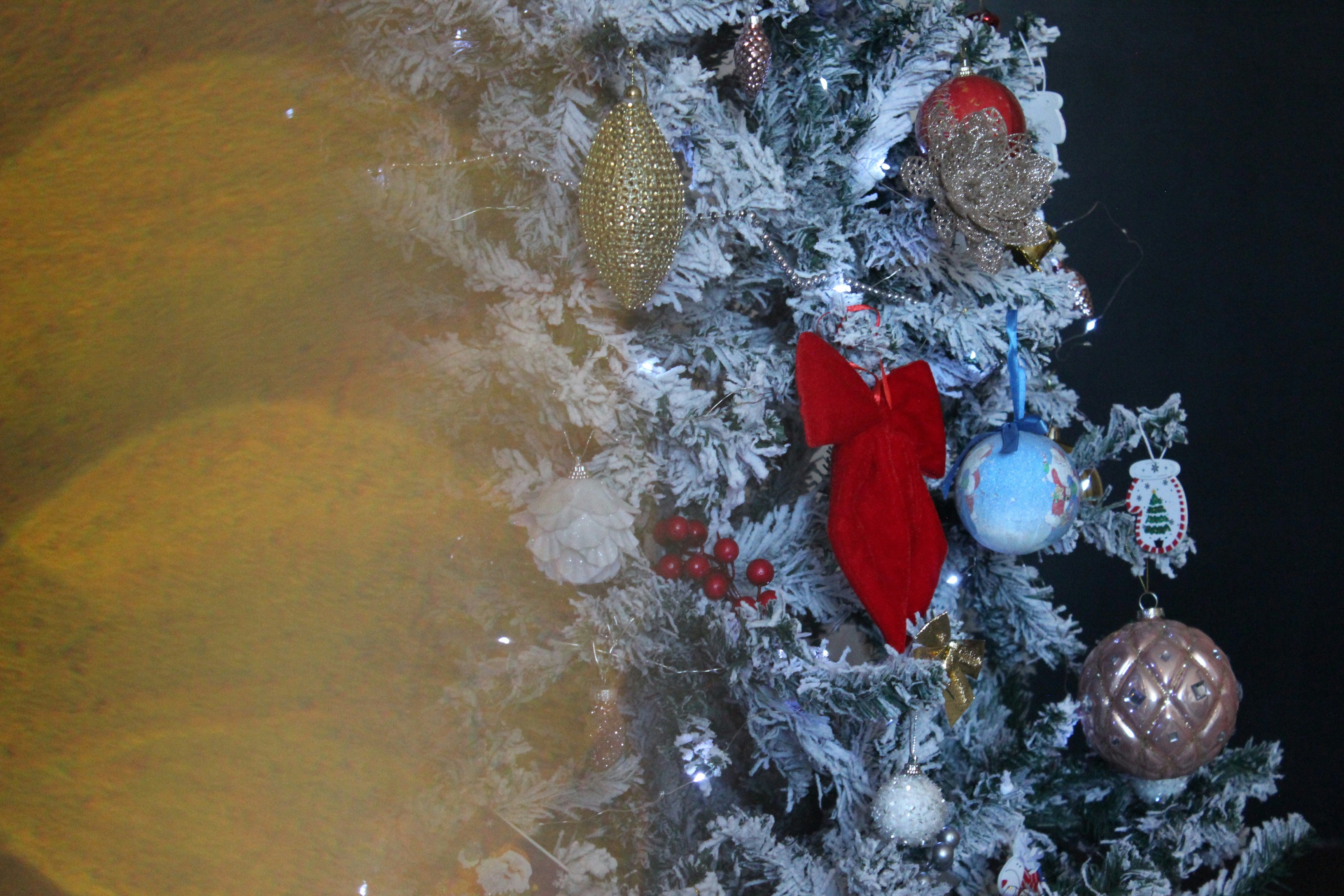 Snow-dusted Christmas tree adorned with red bow and retro baubles, softly illuminated from the left, suggesting a cozy holiday vignette.