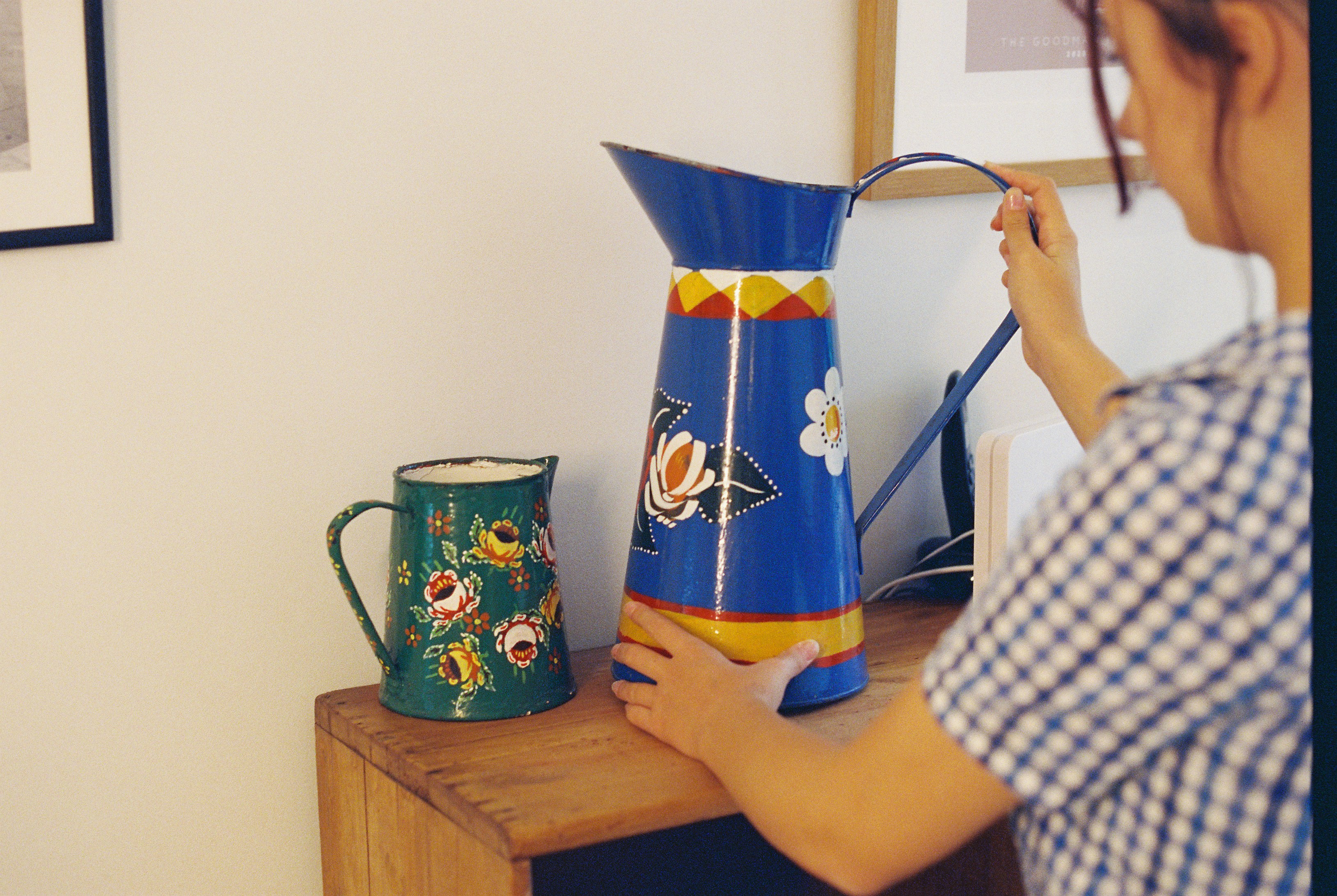 A woman lifts a tall blue folk-painted jug into place beside a smaller green one on a worn pine shelf.