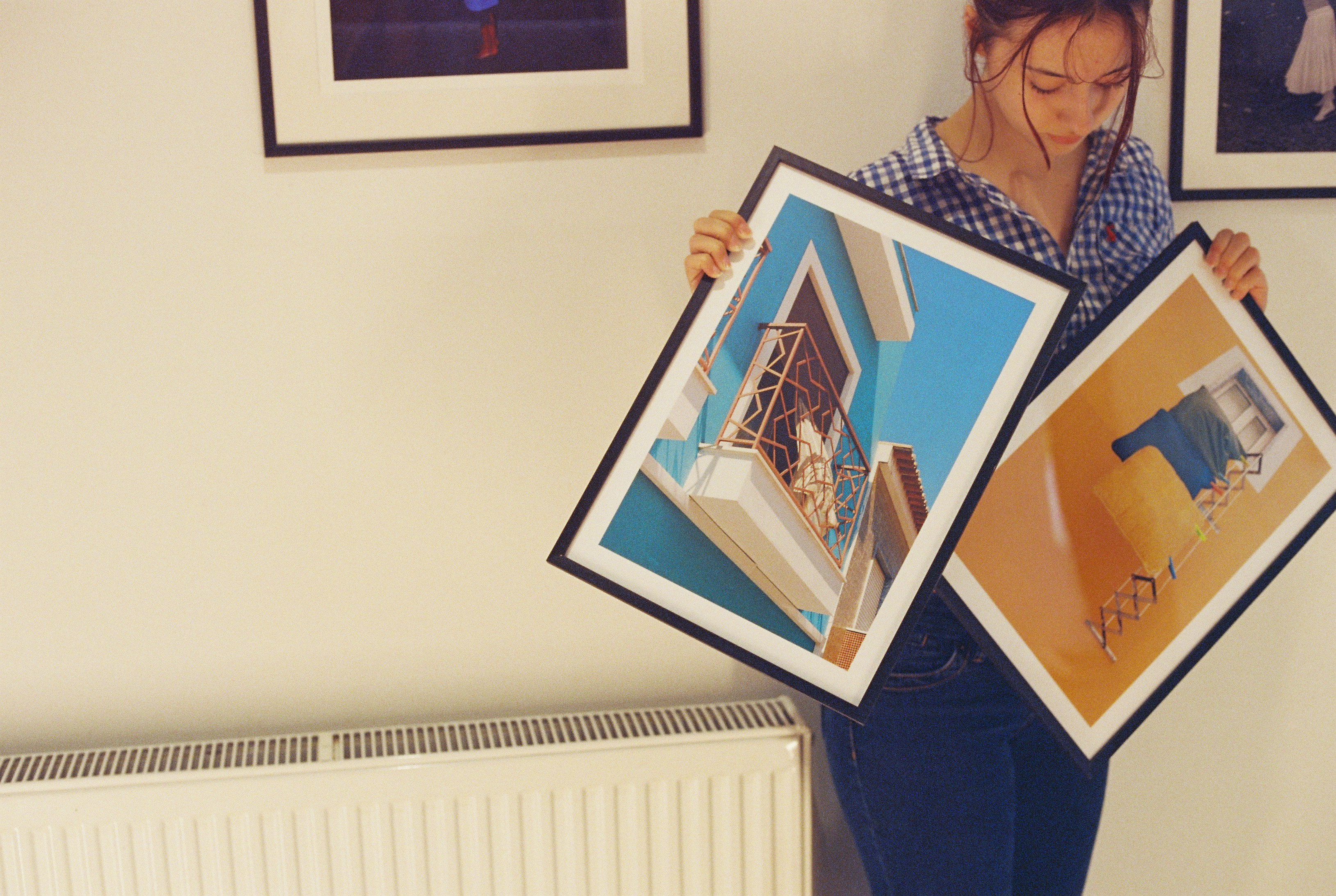 Two framed pictures held out at angles while a woman studies them against the wall she is filling with artwork.
