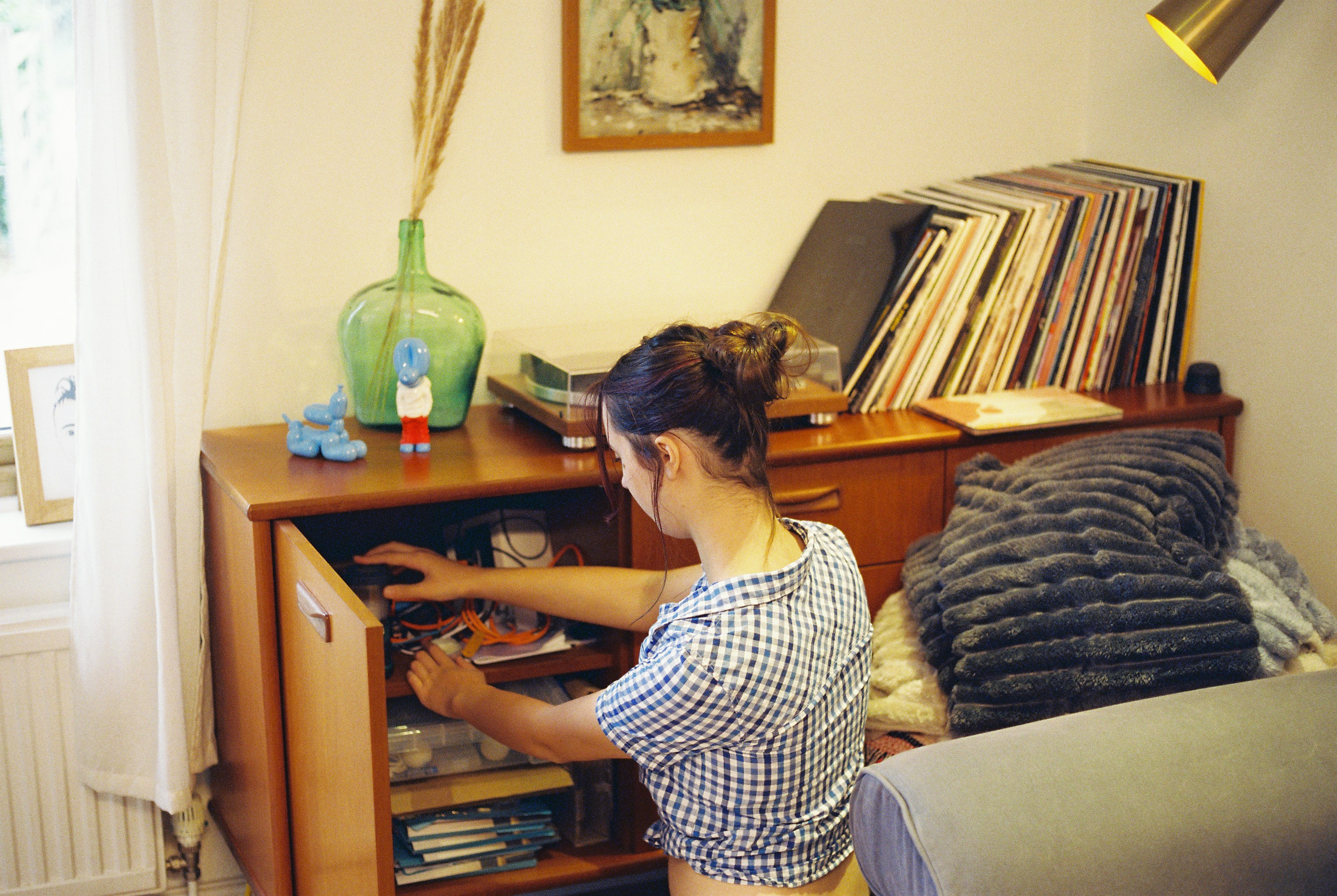 A woman kneels to sort cables and clutter out of a teak cabinet, a row of vinyl records standing above it in soft window light.