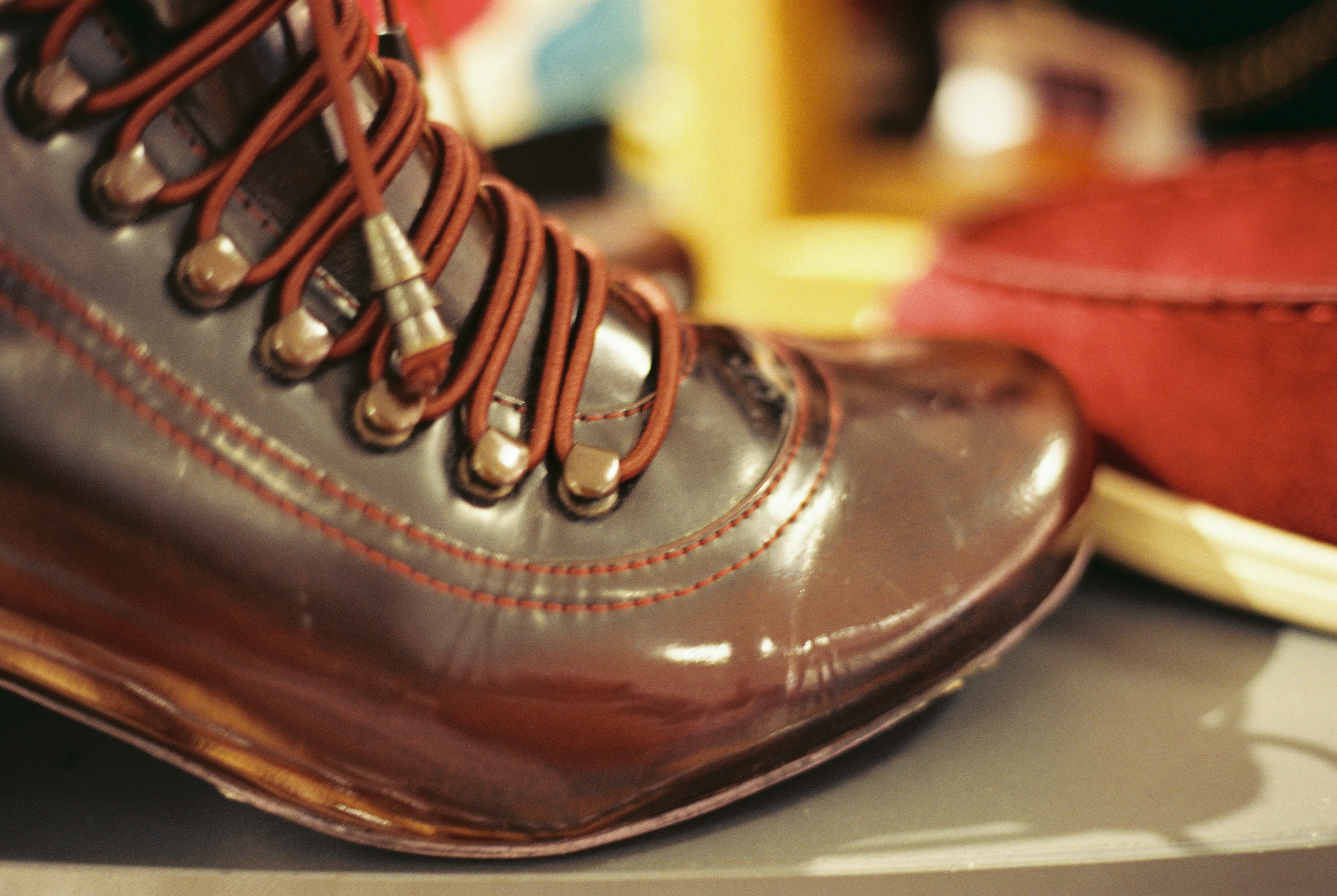 Close-up of a polished brown leather boot with red laces, resting on a reflective surface with warm background tones; indicates merchandise or footwear display.
