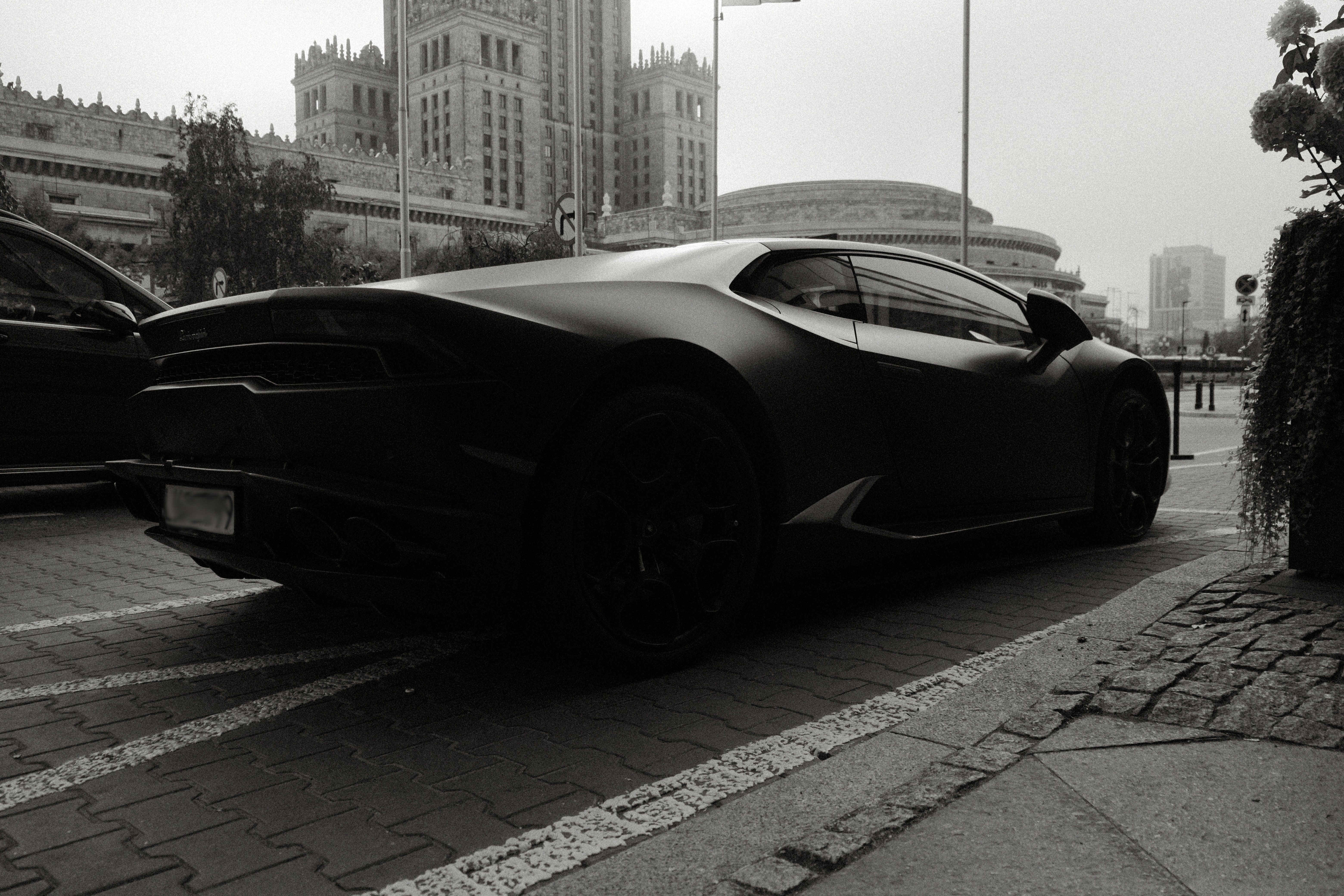 A sleek black sports car parked along a cobblestone street with a cityscape background in grayscale. The emphasis is on the car's silhouette and urban setting.