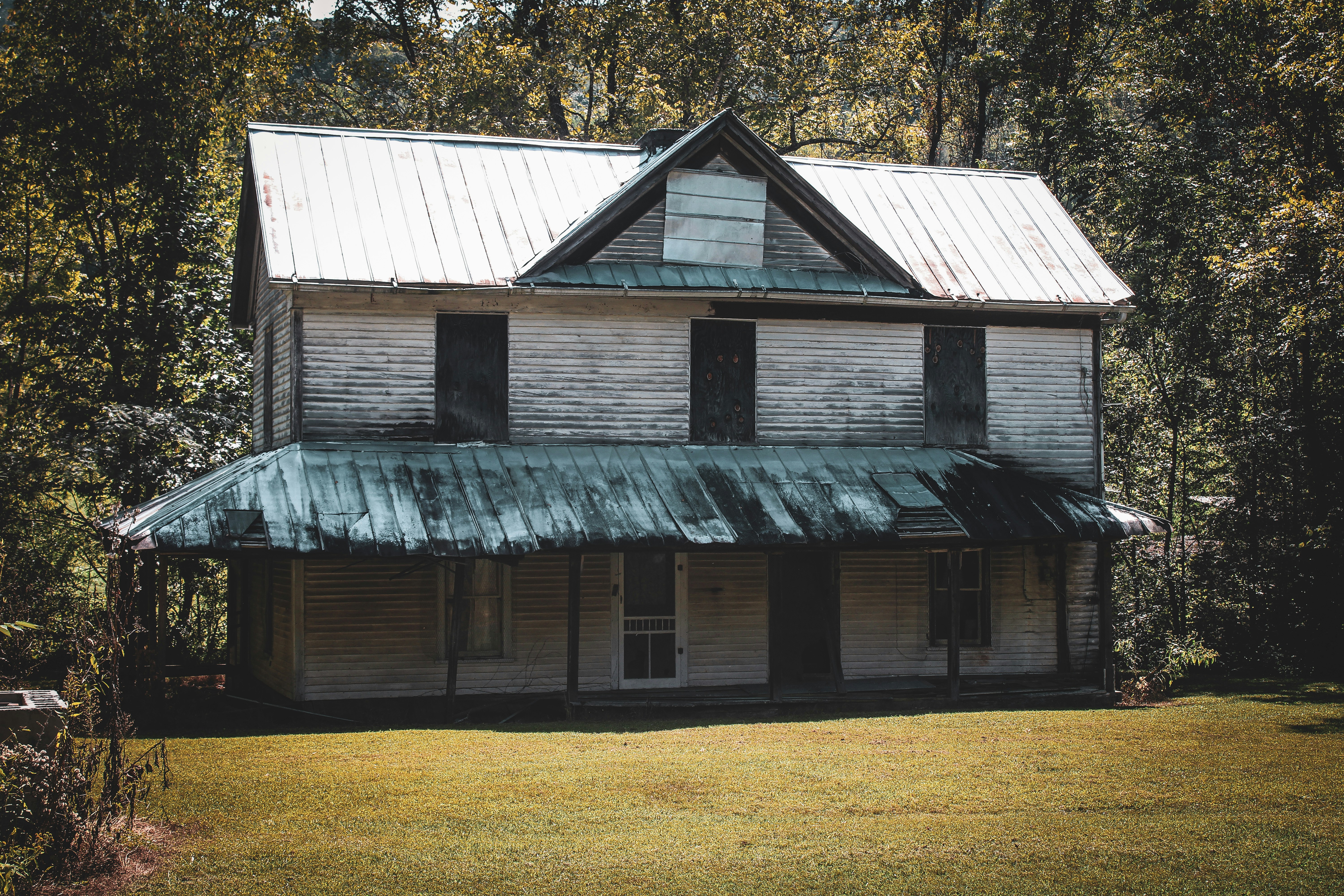 An abandoned two-story white farmhouse with a metal roof in a wooded clearing
