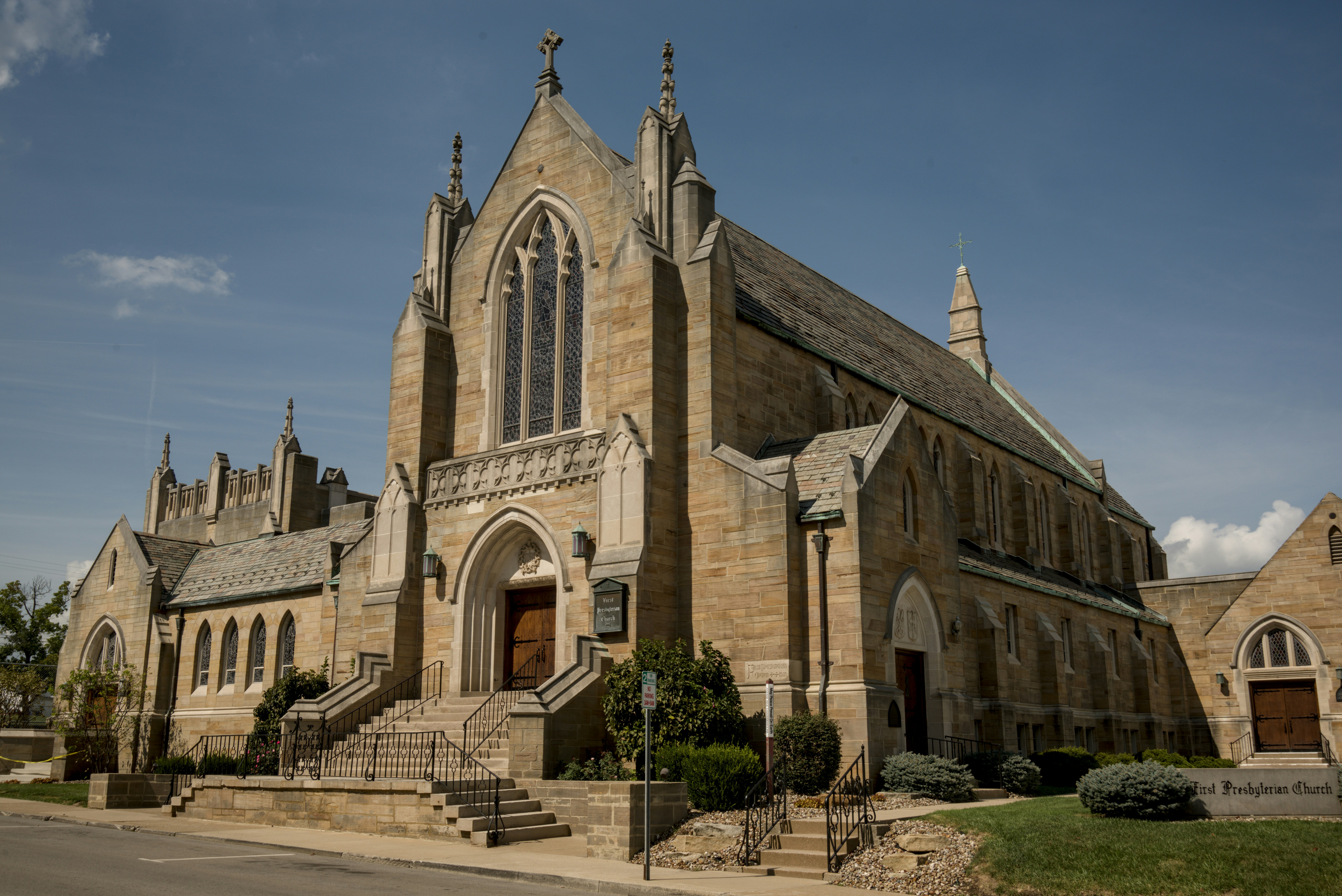 A grand stone church with tall arched windows and steps leading to the entrance, captured under a bright blue sky. The scene emphasizes architectural detail and surrounding landscape.