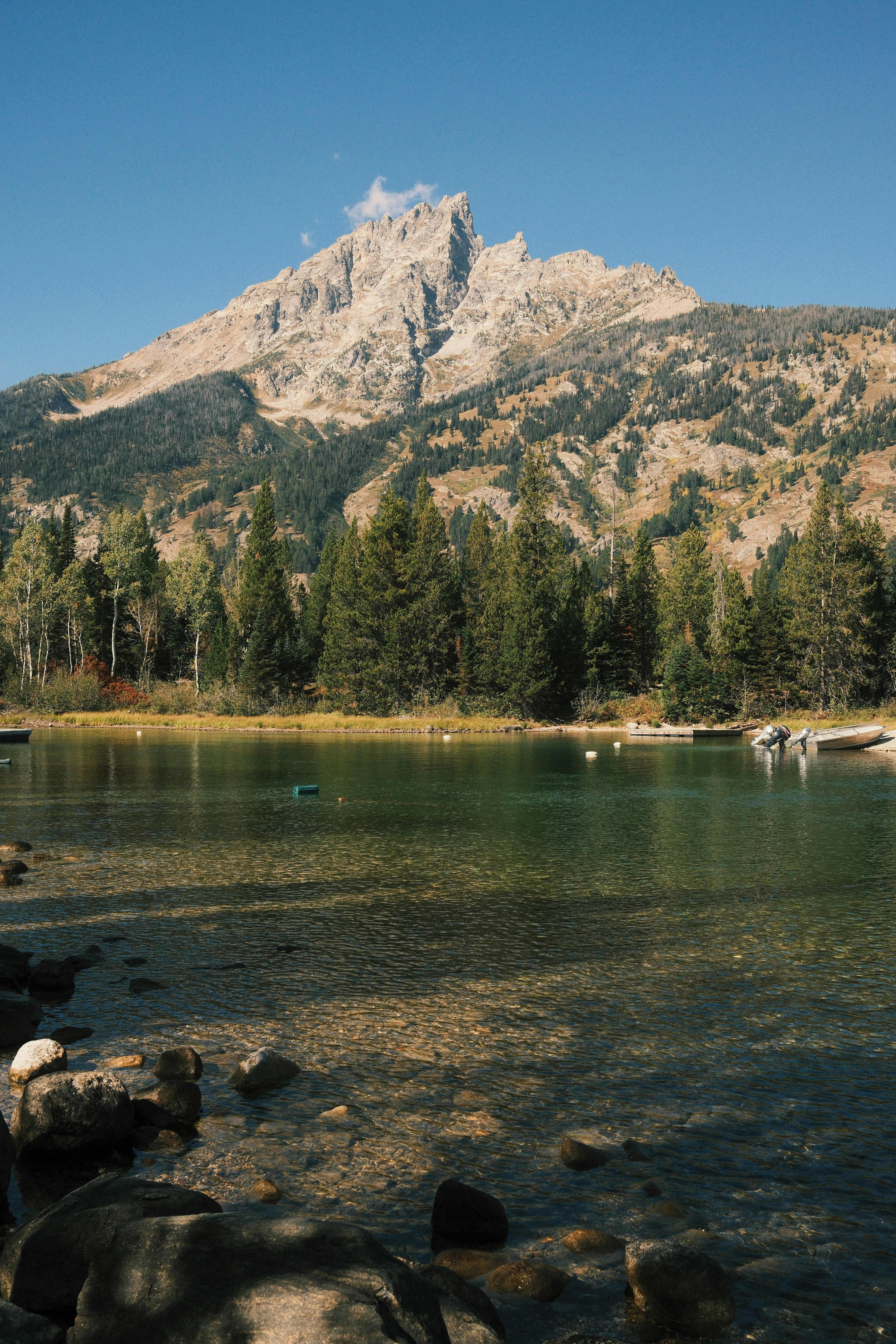 Clear alpine lake at the base of rugged peaks with a forested shoreline and rocky foreground.