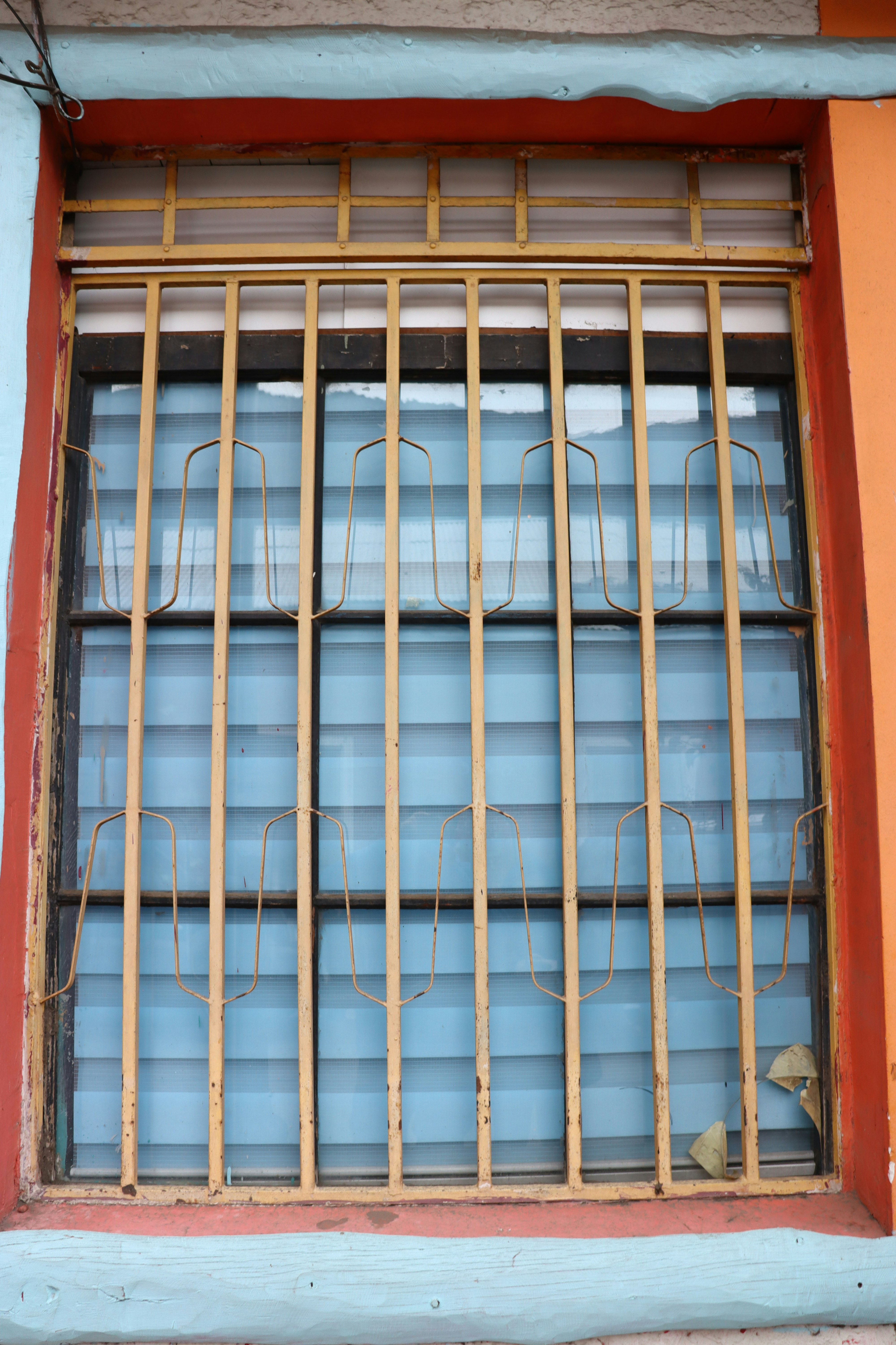 Vertical metal bars guard a blue shuttered window framed by orange masonry, with a pale aqua sill and a pale blue bottom trim.