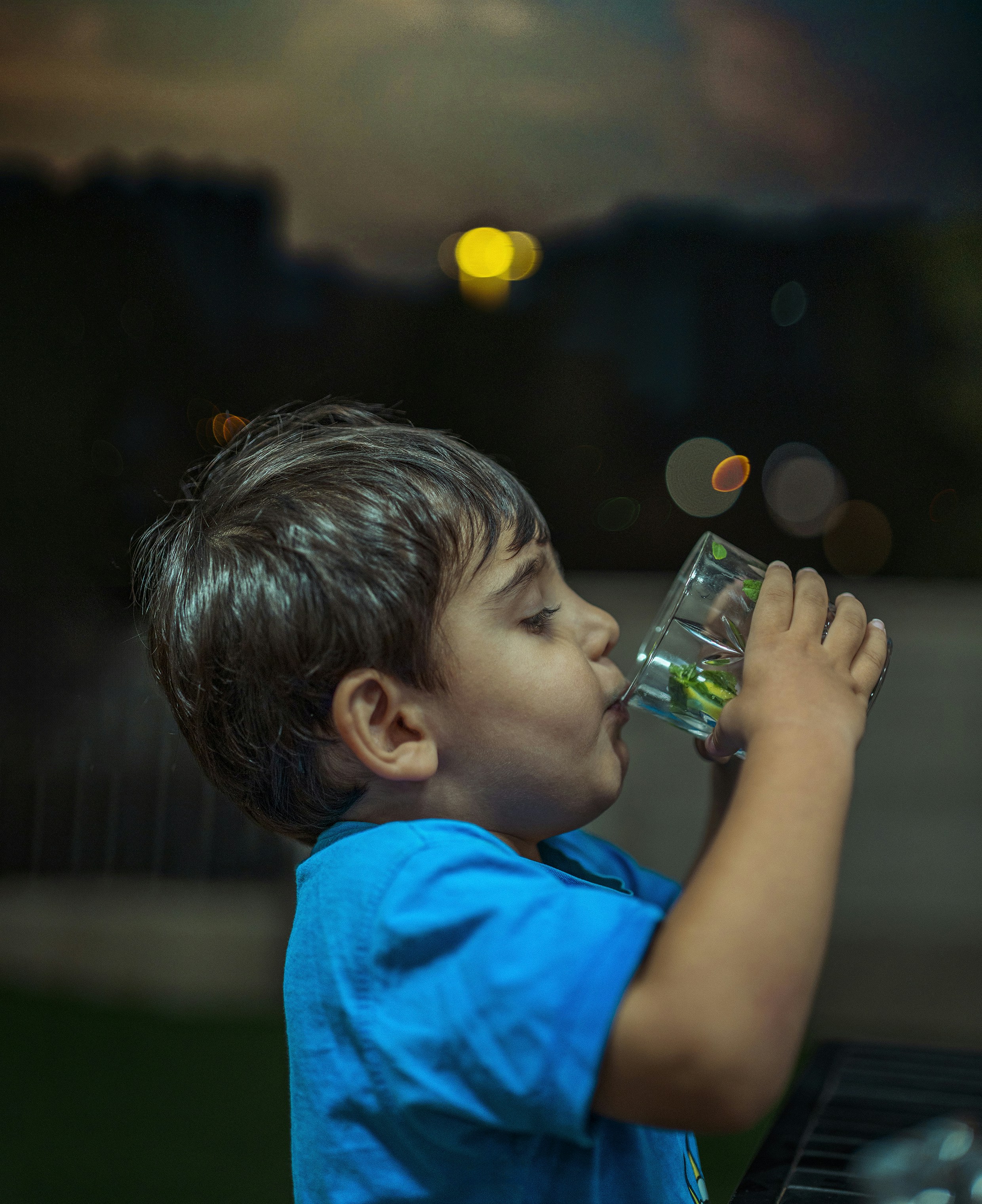 Young boy in a bright blue shirt drinks from a glass at dusk, the background softly blurred with bokeh; this is a candid portrait capturing a simple moment.