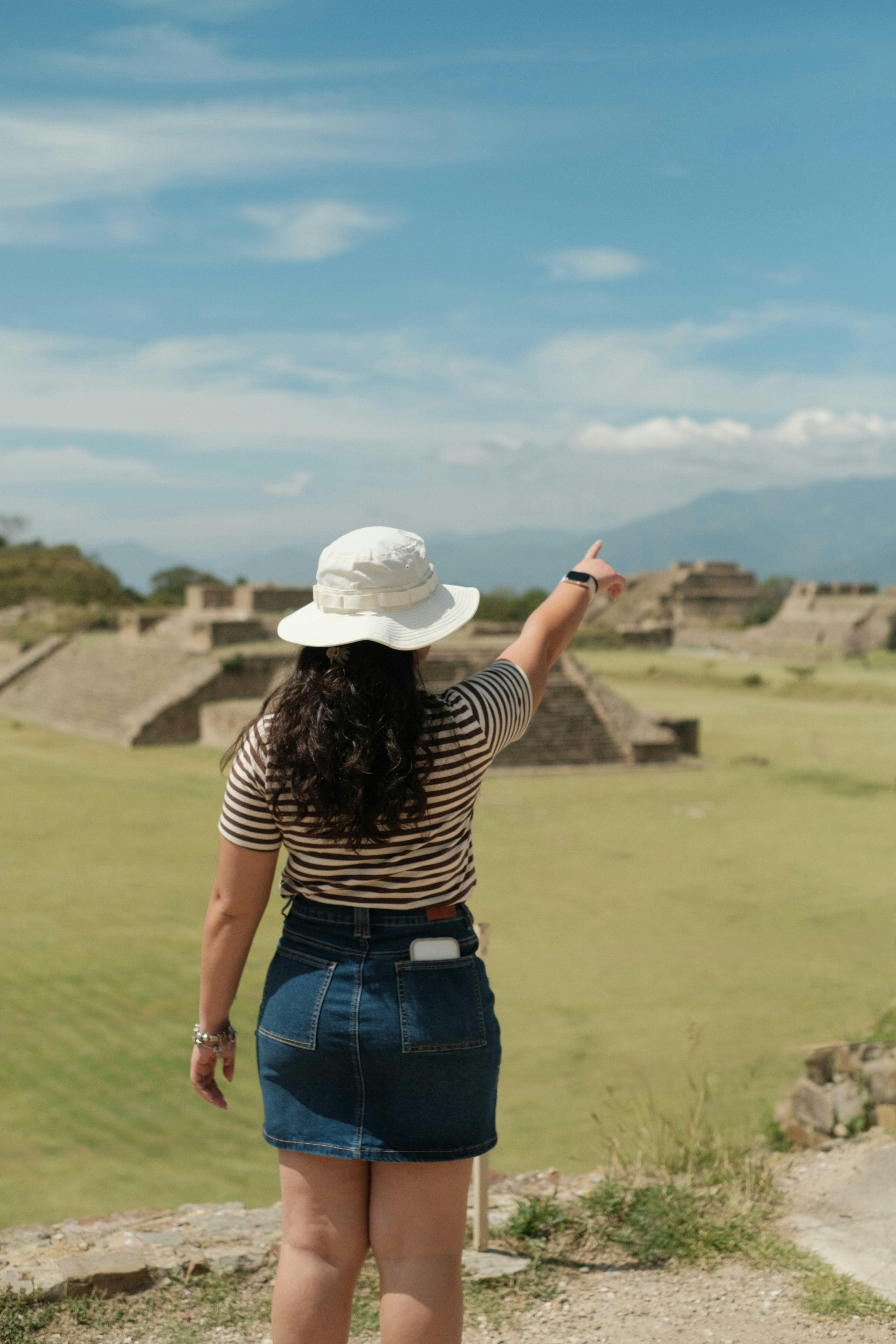 A woman with a striped shirt and white hat stands with her back to the camera, pointing toward ancient terraces under a blue sky.