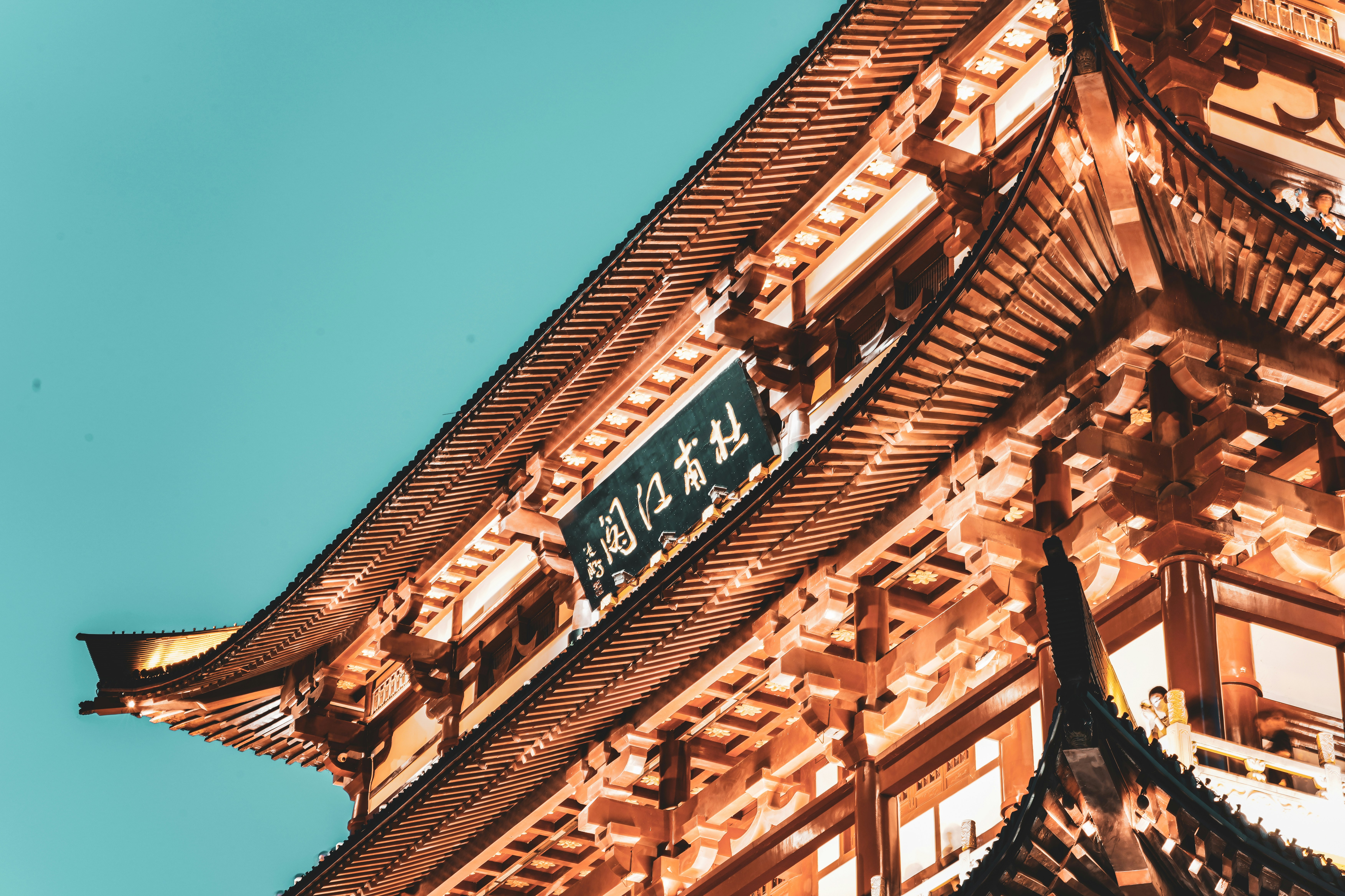 A close-up architectural shot of an ornate traditional roof with warm lighting against a turquoise sky. The image emphasizes the intricate eaves and latticework of the building, highlighting its cultural detail.