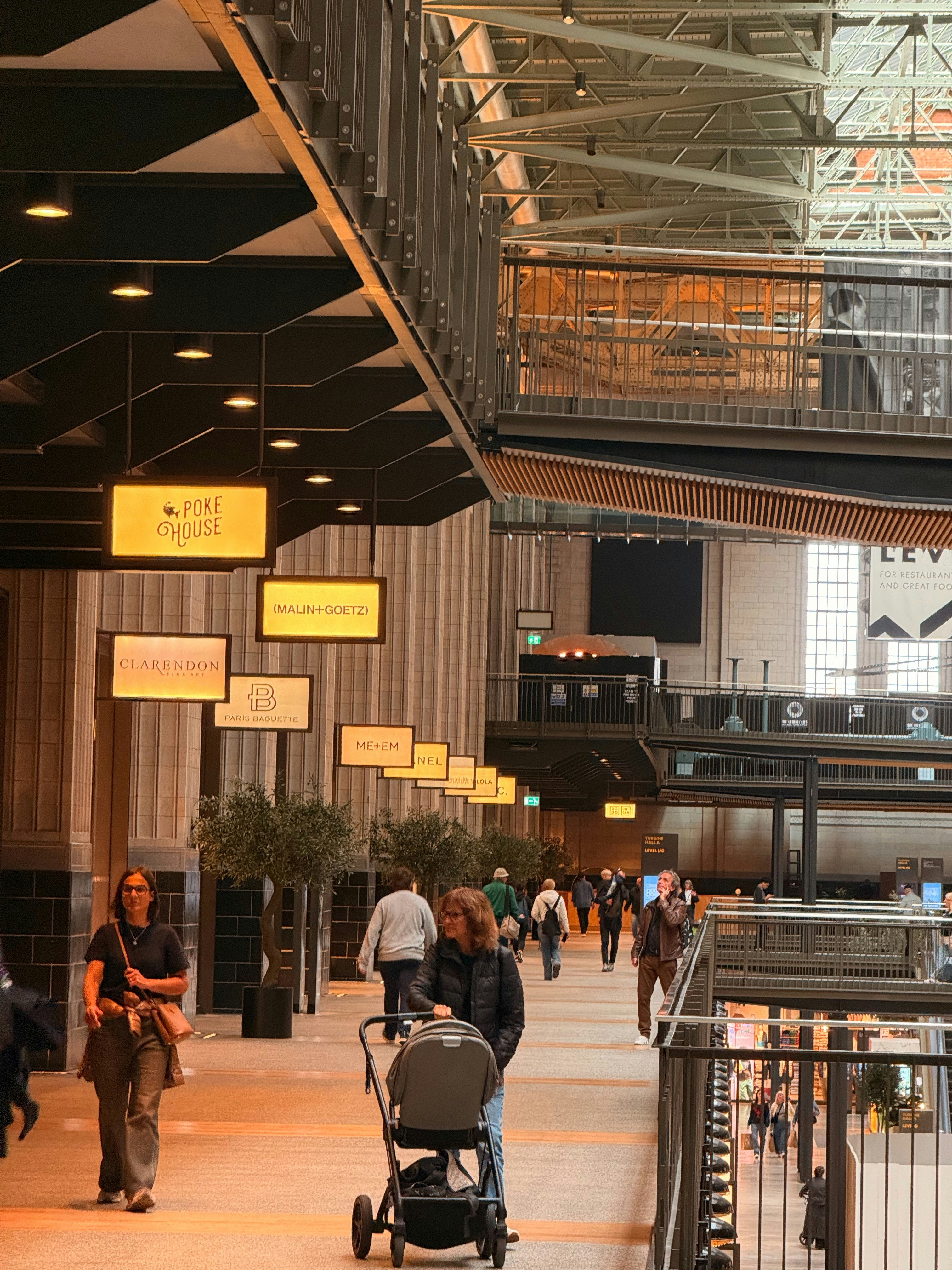 Strolling Through a Sunlit Atrium: A Stroller and Shoppers Beneath Overhead Walkways